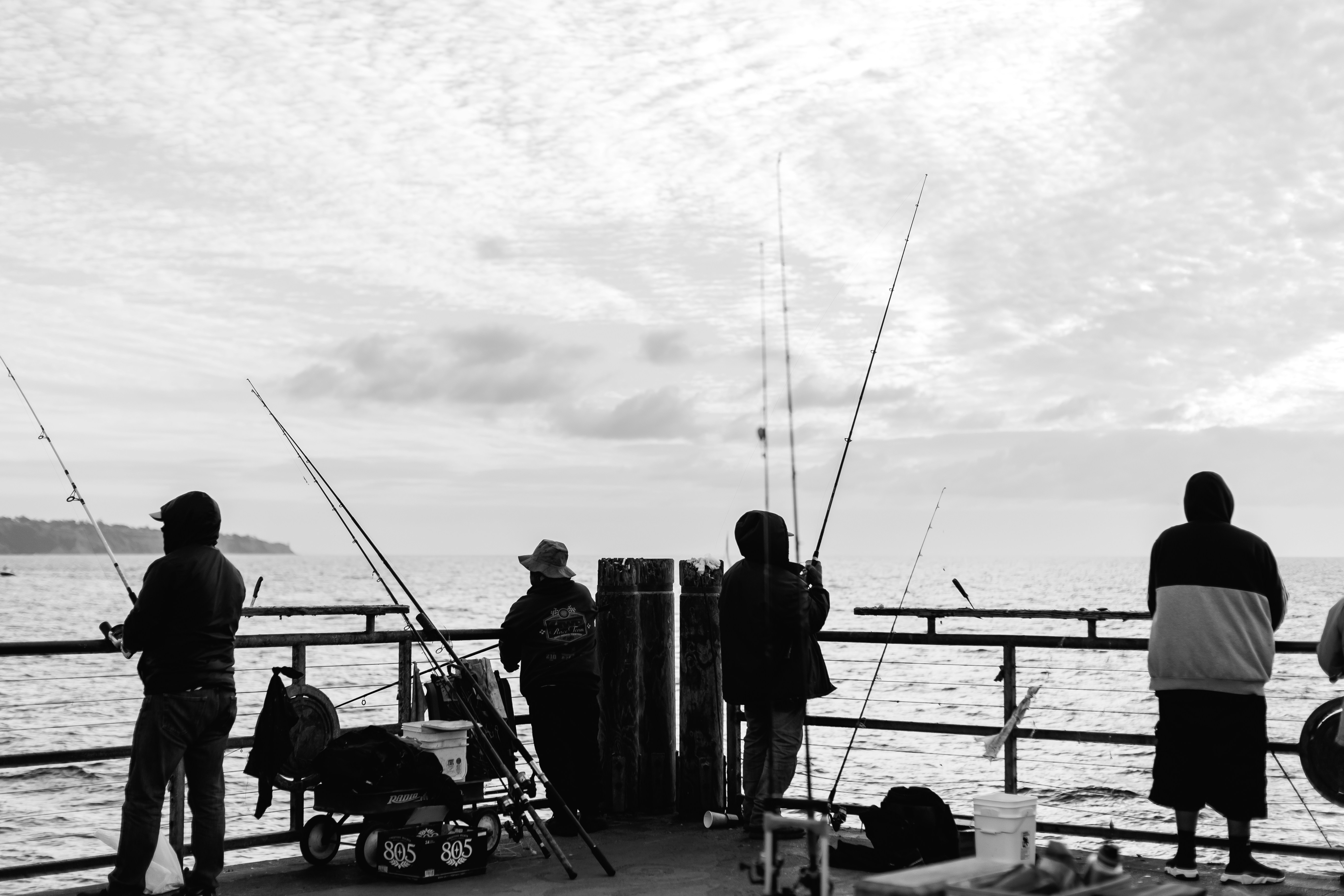 Silhouetted figures fish along a pier under a textured sky in black and white.