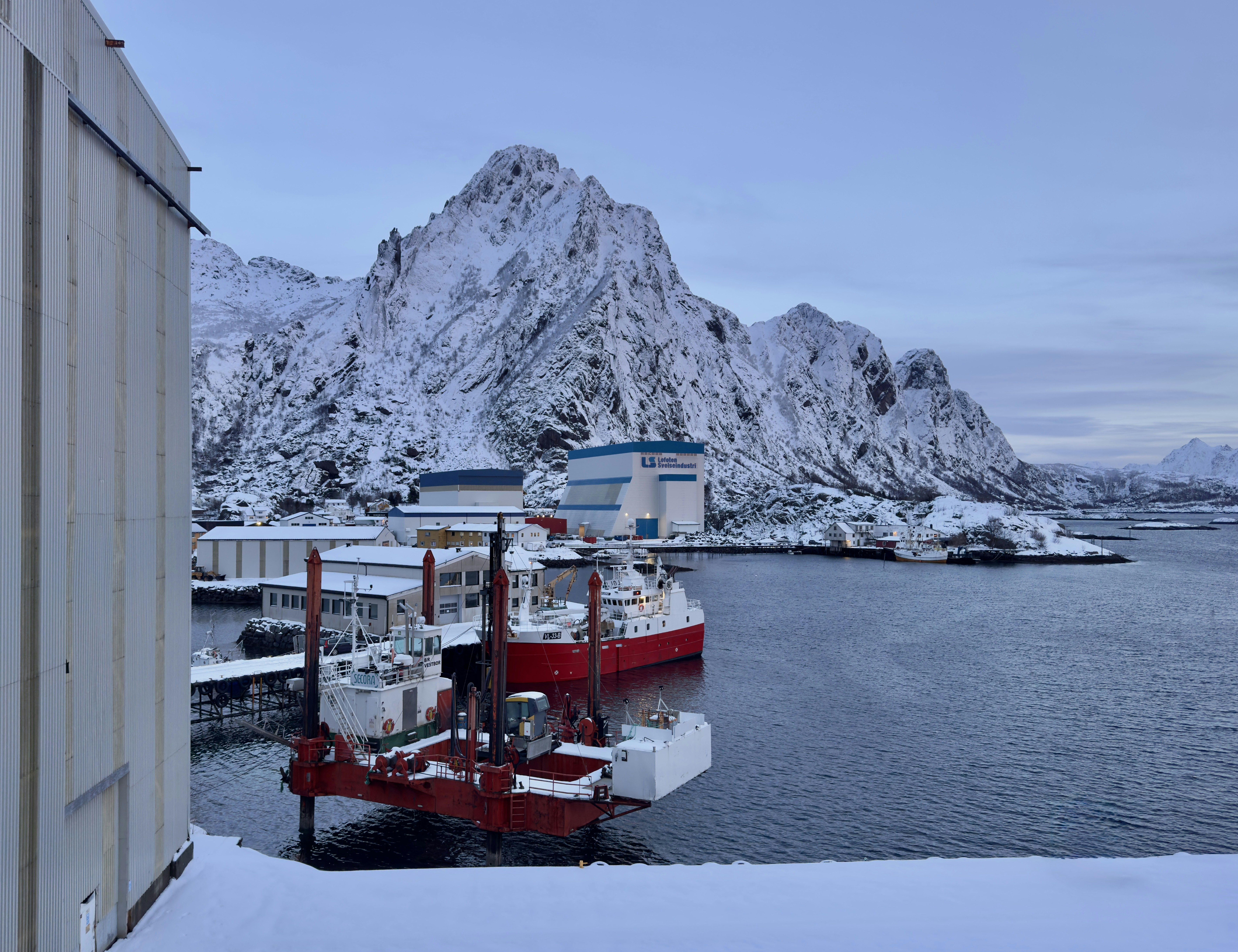 Snow-covered mountains tower over a tranquil harbor with red and white boats docked under a pale blue sky.