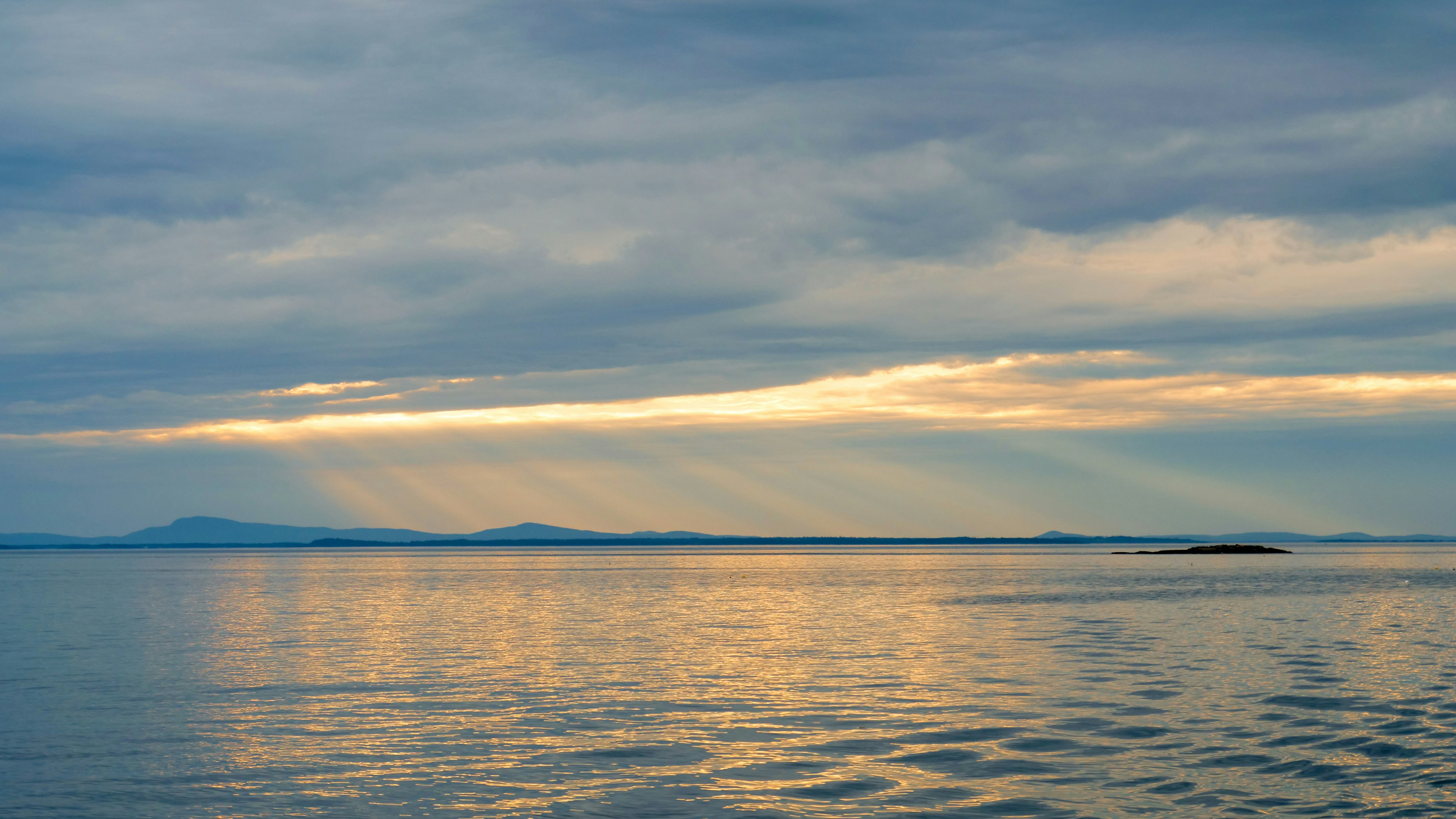 Sunbeams pierce through thick clouds, illuminating a calm sea with golden hues.