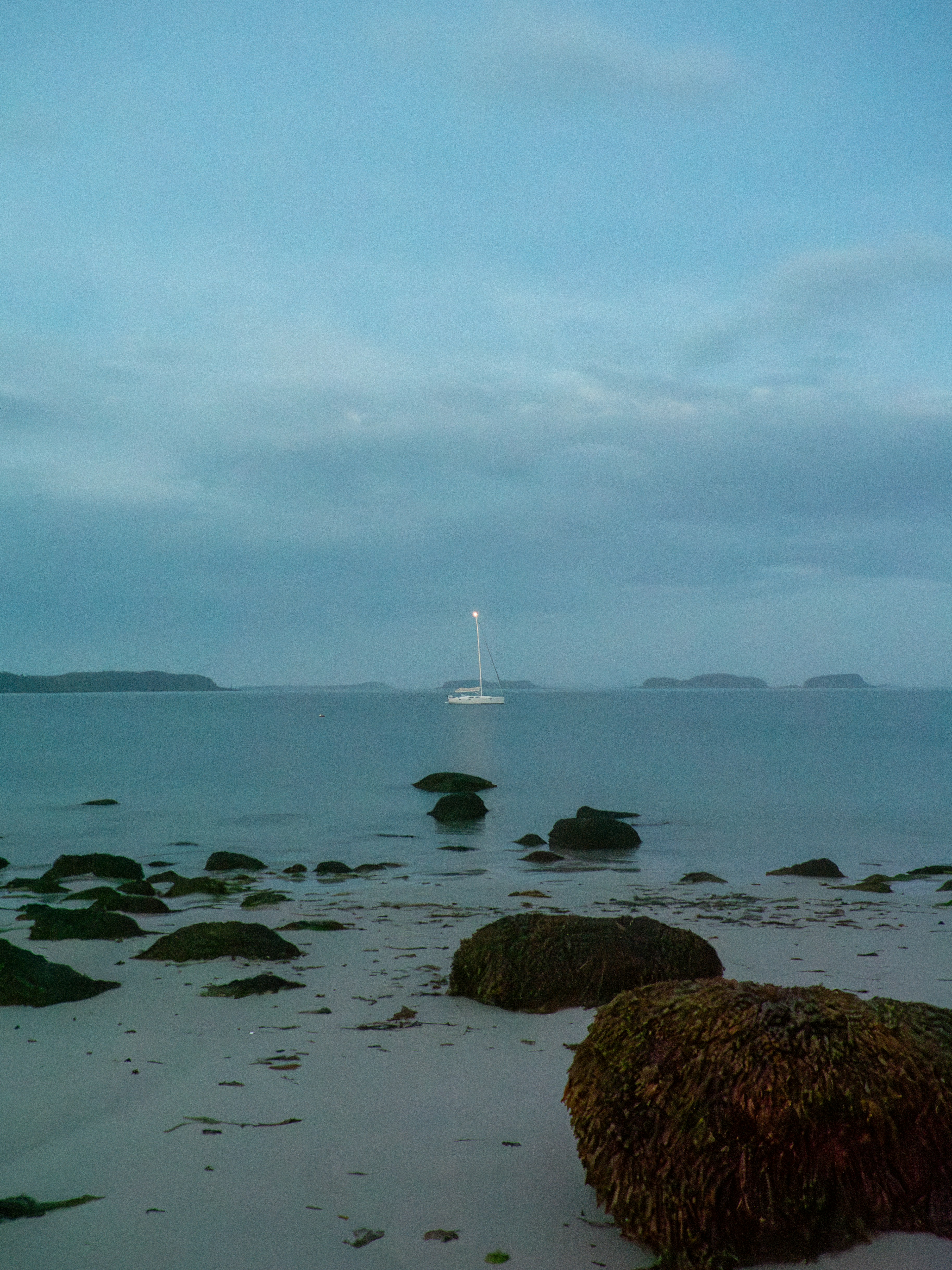 Sailboat anchored near a rocky shoreline under an overcast sky.
