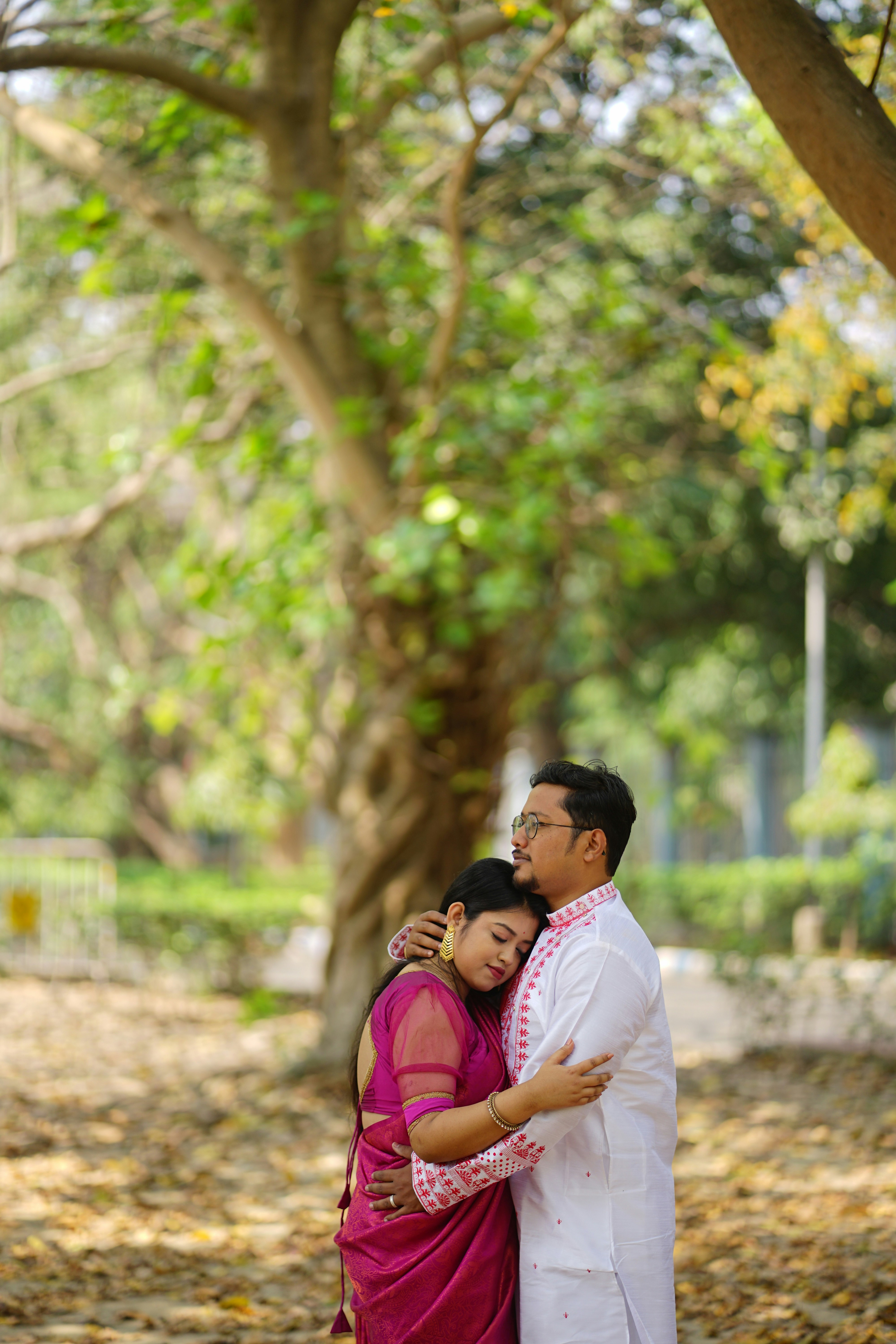 Couple embracing beneath a large tree in a sunlit park with fallen leaves.
