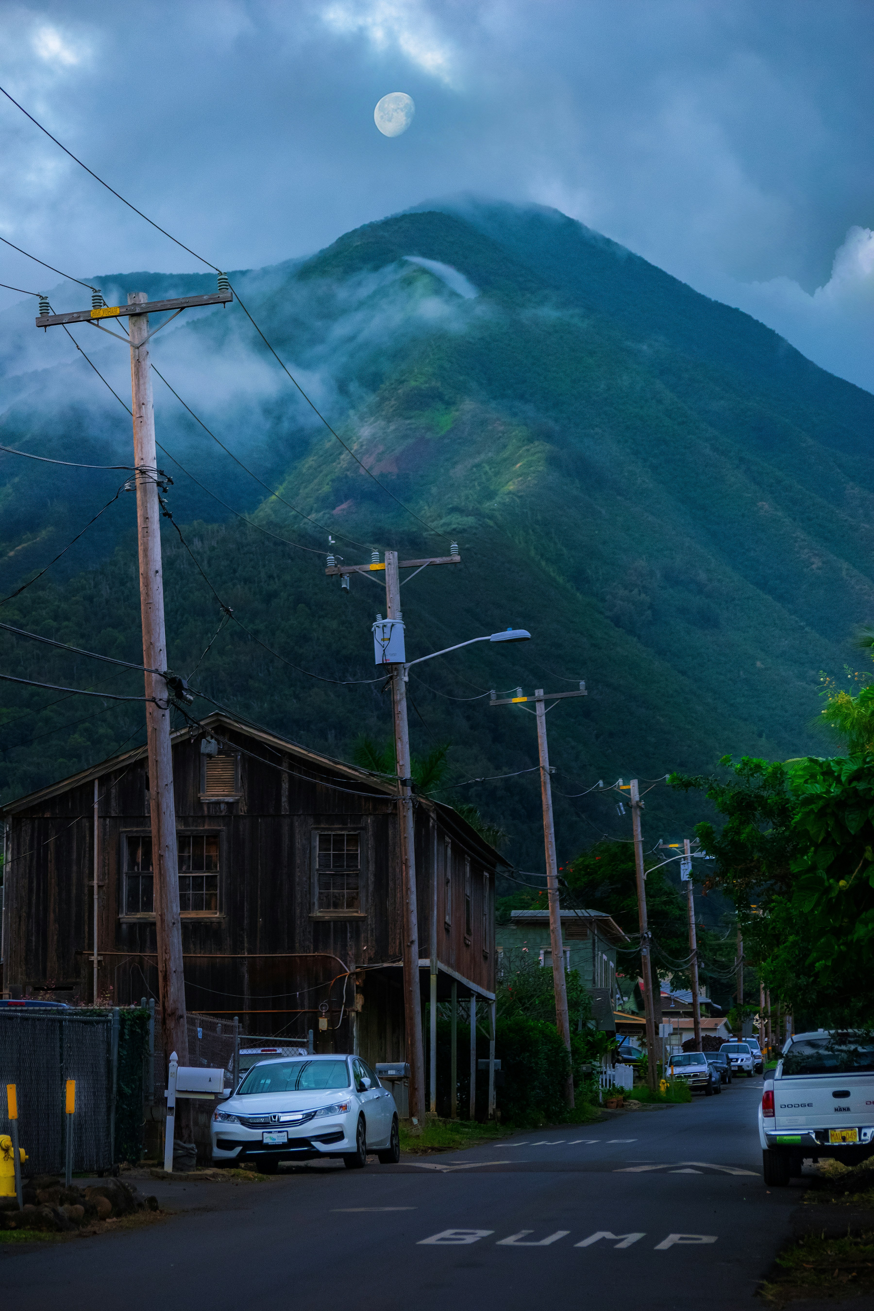 Quiet street with a rustic building under a misty mountain and a visible moon in the evening sky.