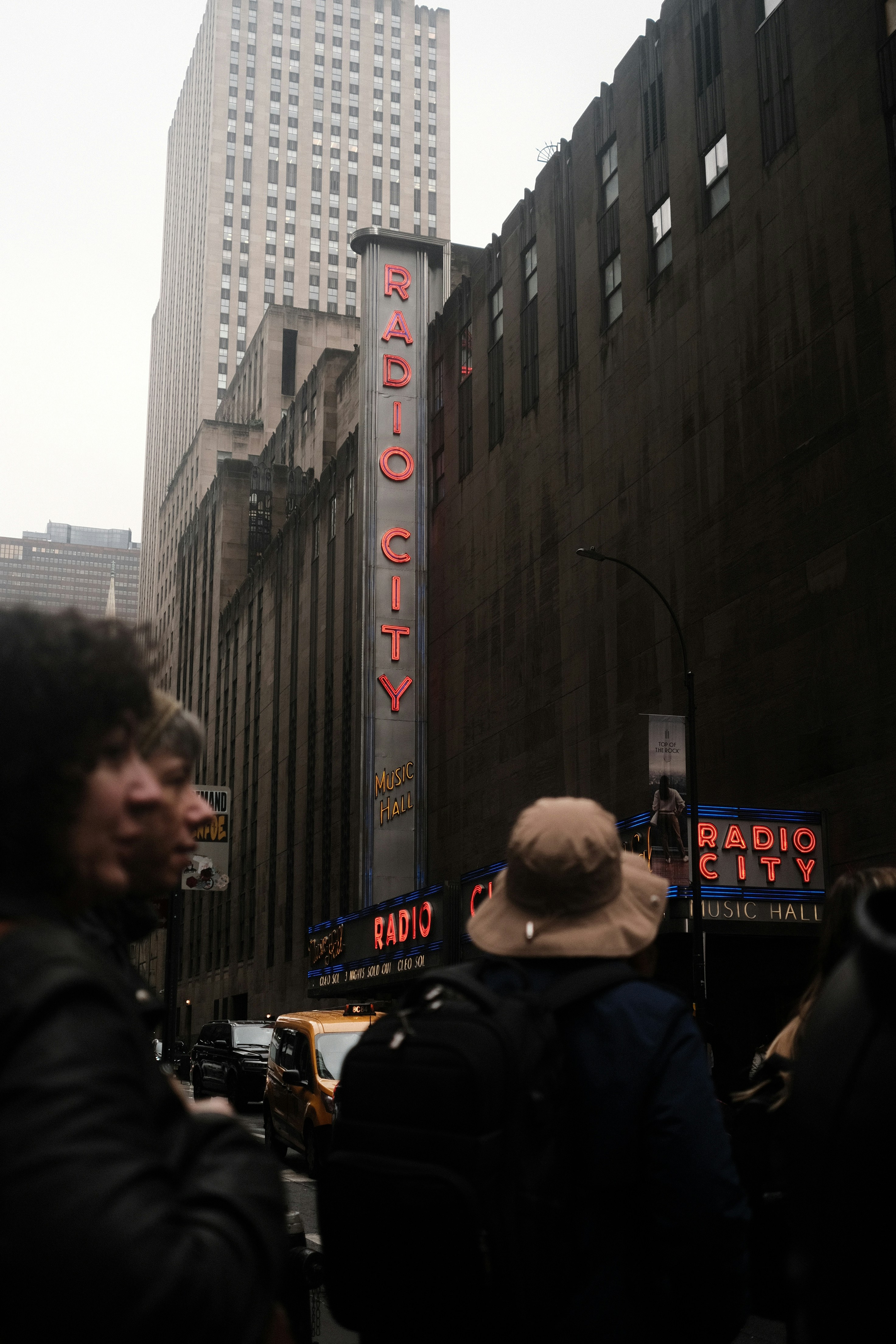 Radio City Music Hall's iconic neon sign against a towering cityscape with pedestrians in the foreground.