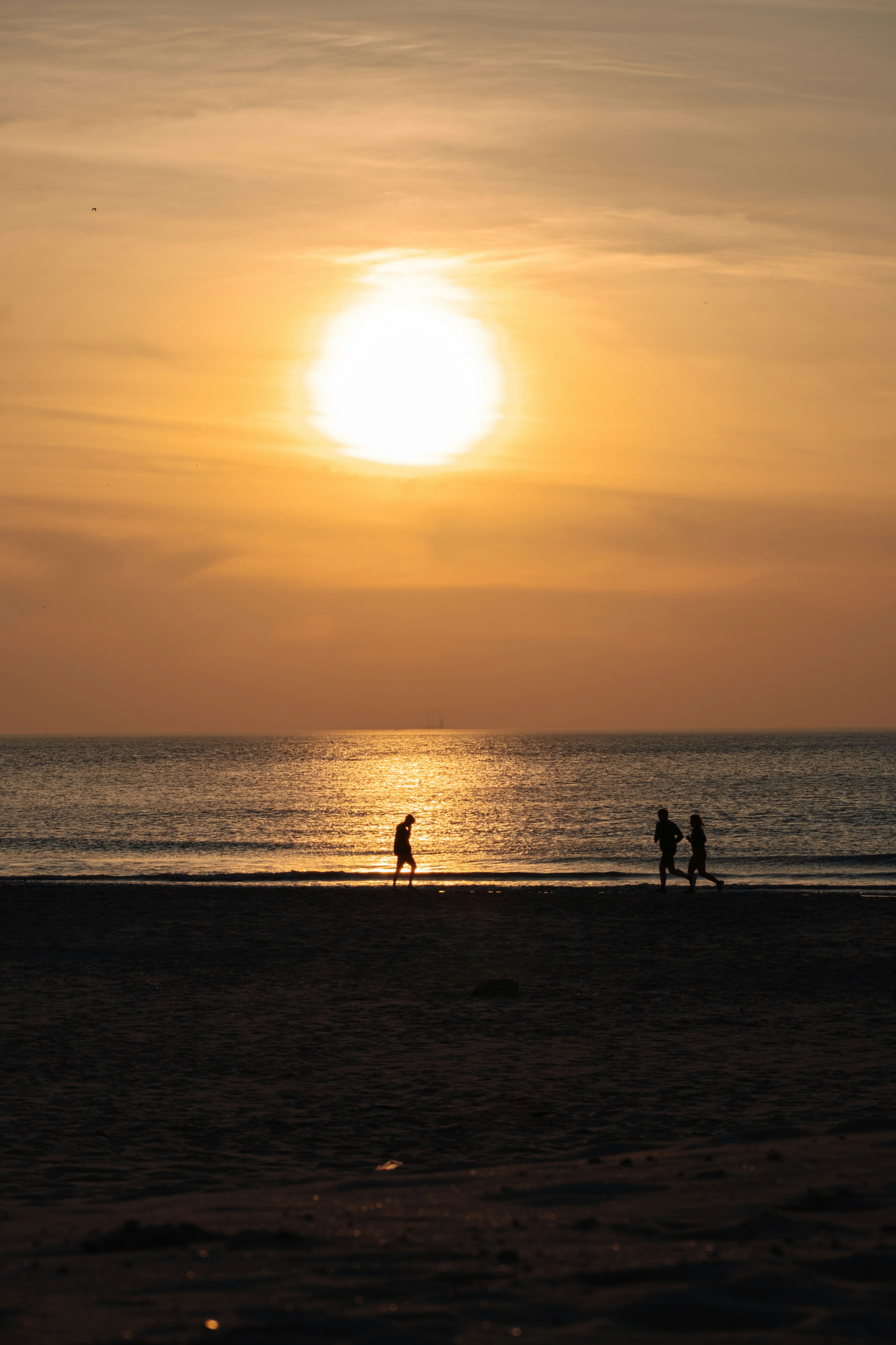 Silhouetted figures walk along the shore at sunset with the sun casting a warm glow over the ocean.