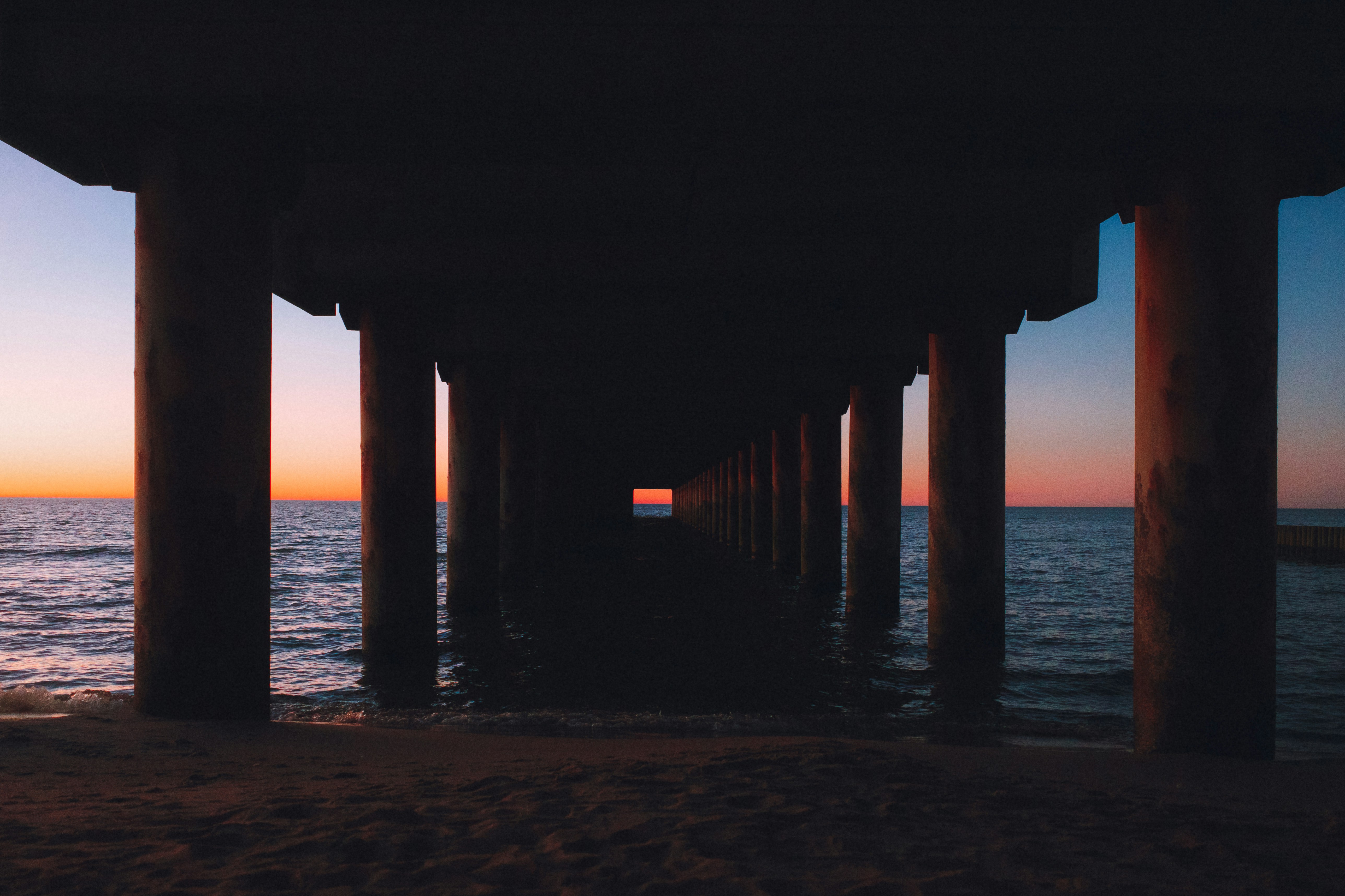 Sunset view from beneath a pier, with silhouetted pillars framing the glowing horizon over the ocean.