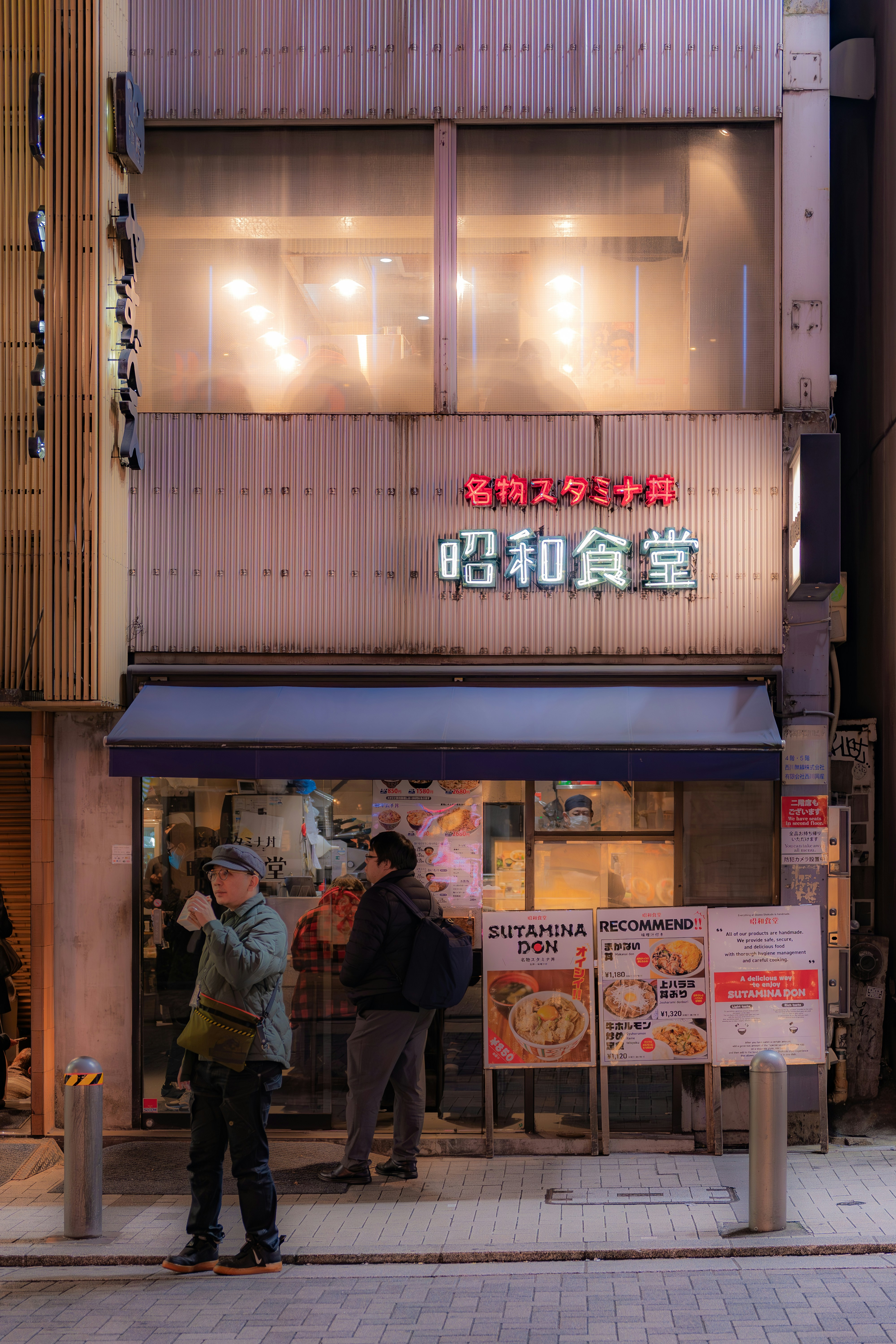 Two people stand outside a warmly lit restaurant on a city street at dusk.