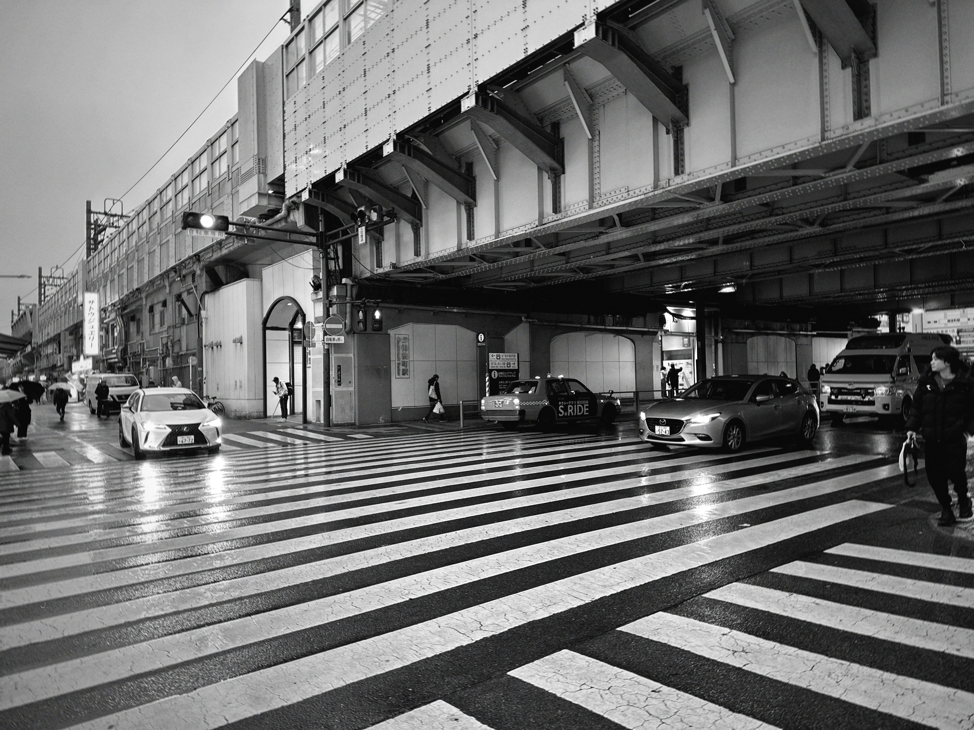 Monochrome view of a rainy Tokyo street with cars and pedestrians at a crosswalk under an overpass.