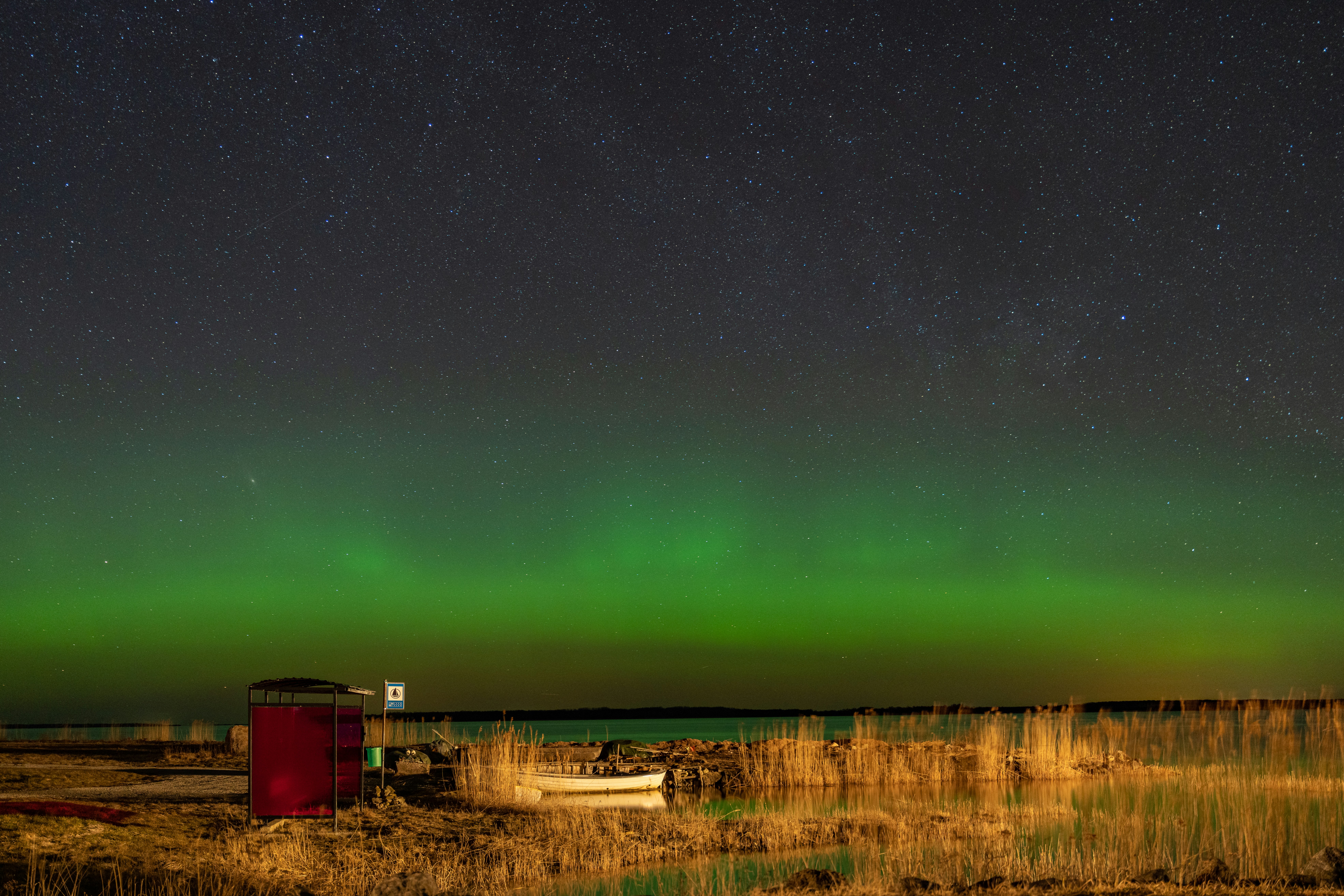 Northern lights illuminate a calm shoreline with a rustic shack and boat under a starry sky.
