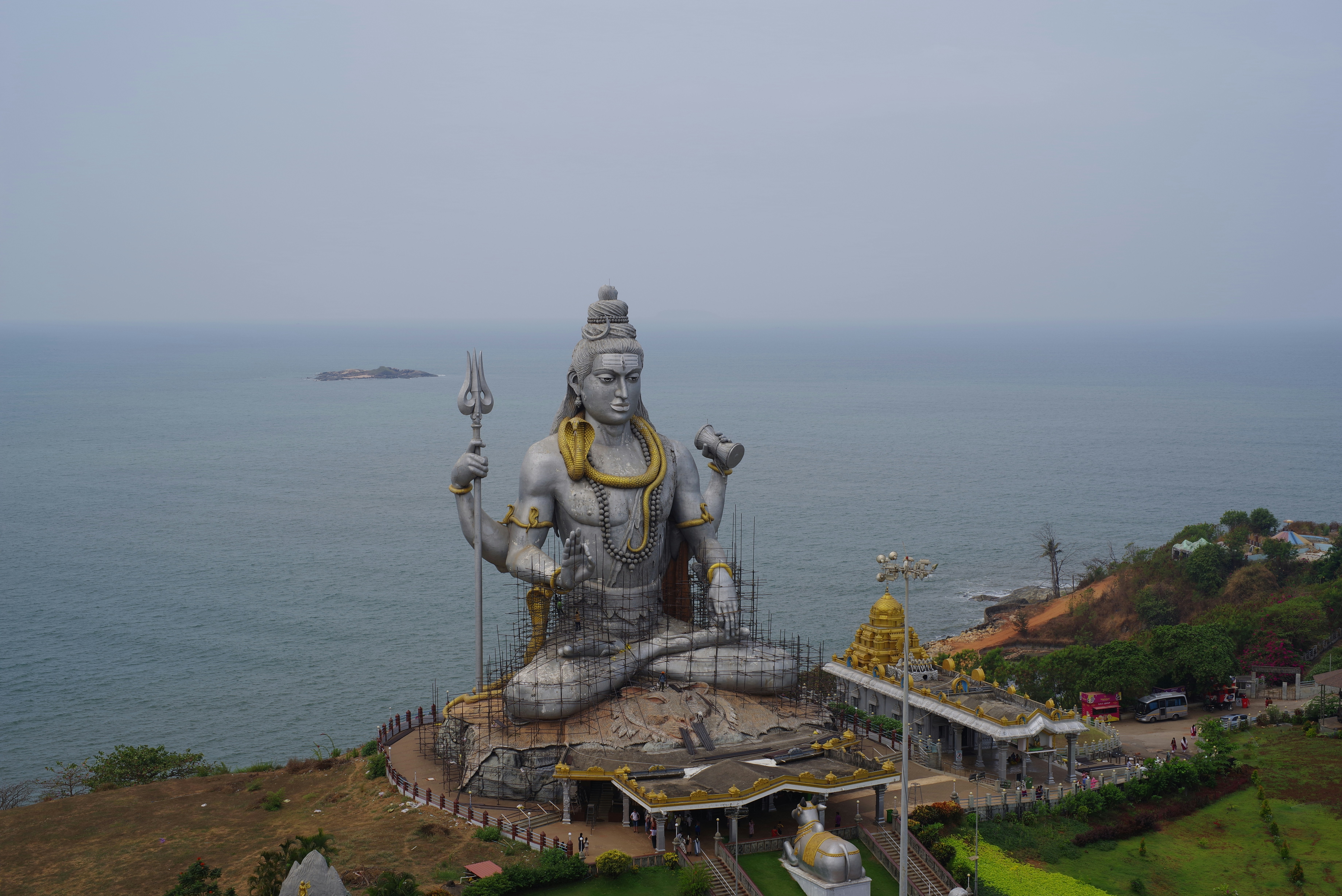 Enormous statue of Lord Shiva seated by the ocean with verdant surroundings under a cloudy sky.