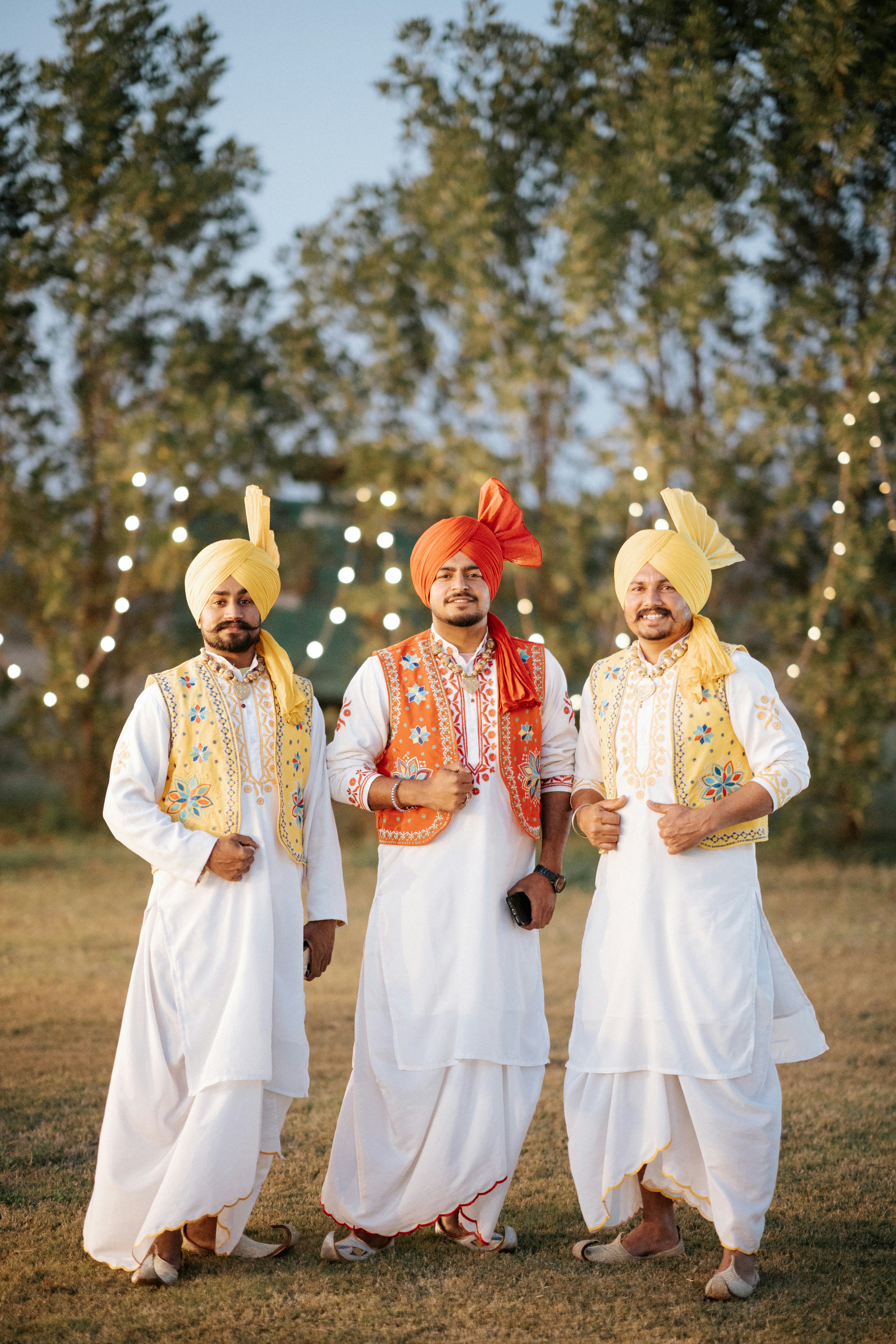 Three men in vibrant traditional attire stand on grass with string lights and trees in the background.