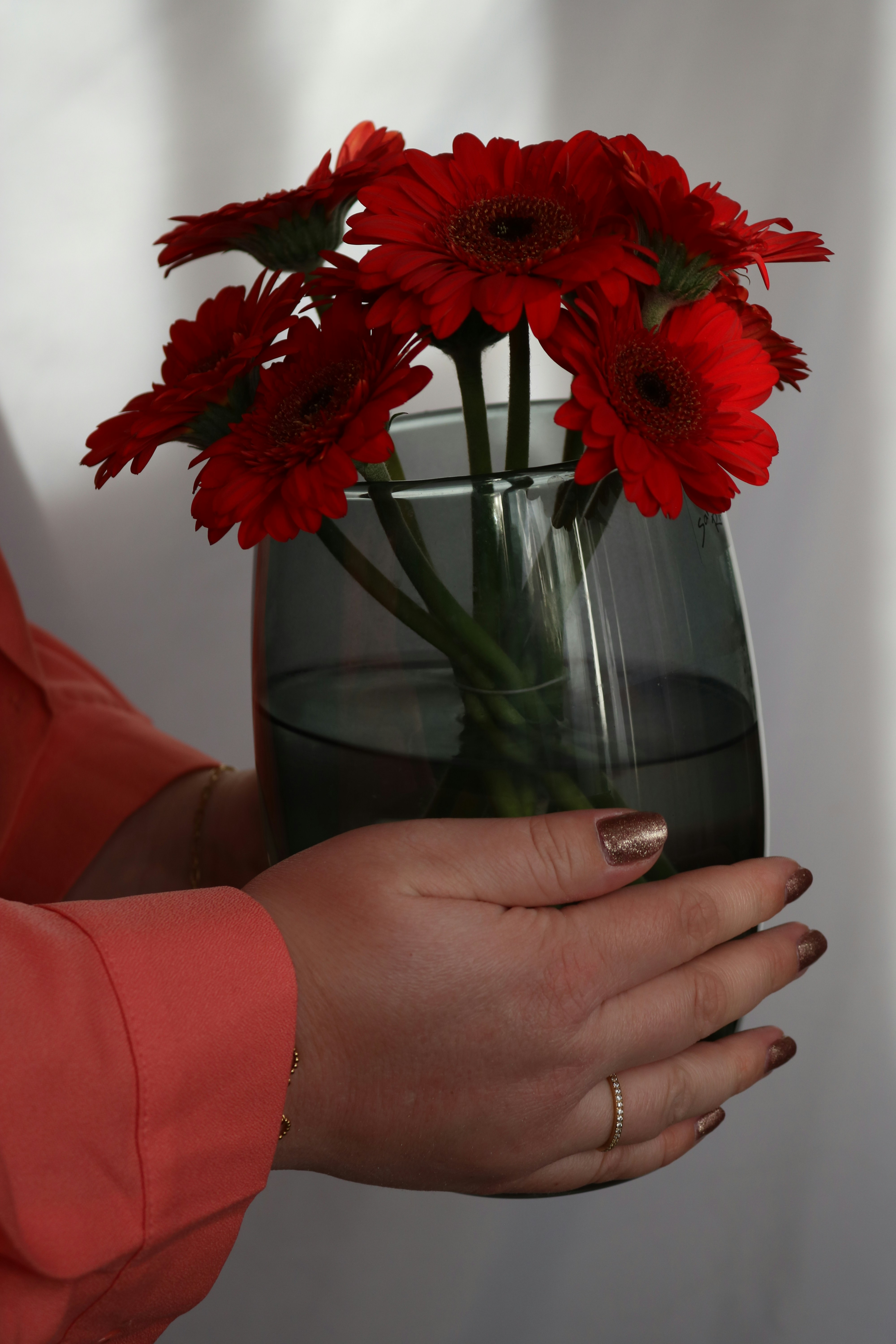 A hand cradles a glass vase filled with bright red gerbera daisies, set against a softly blurred background. The contrast between the flowers and the subtle tones enhances the floral arrangement's vividness.