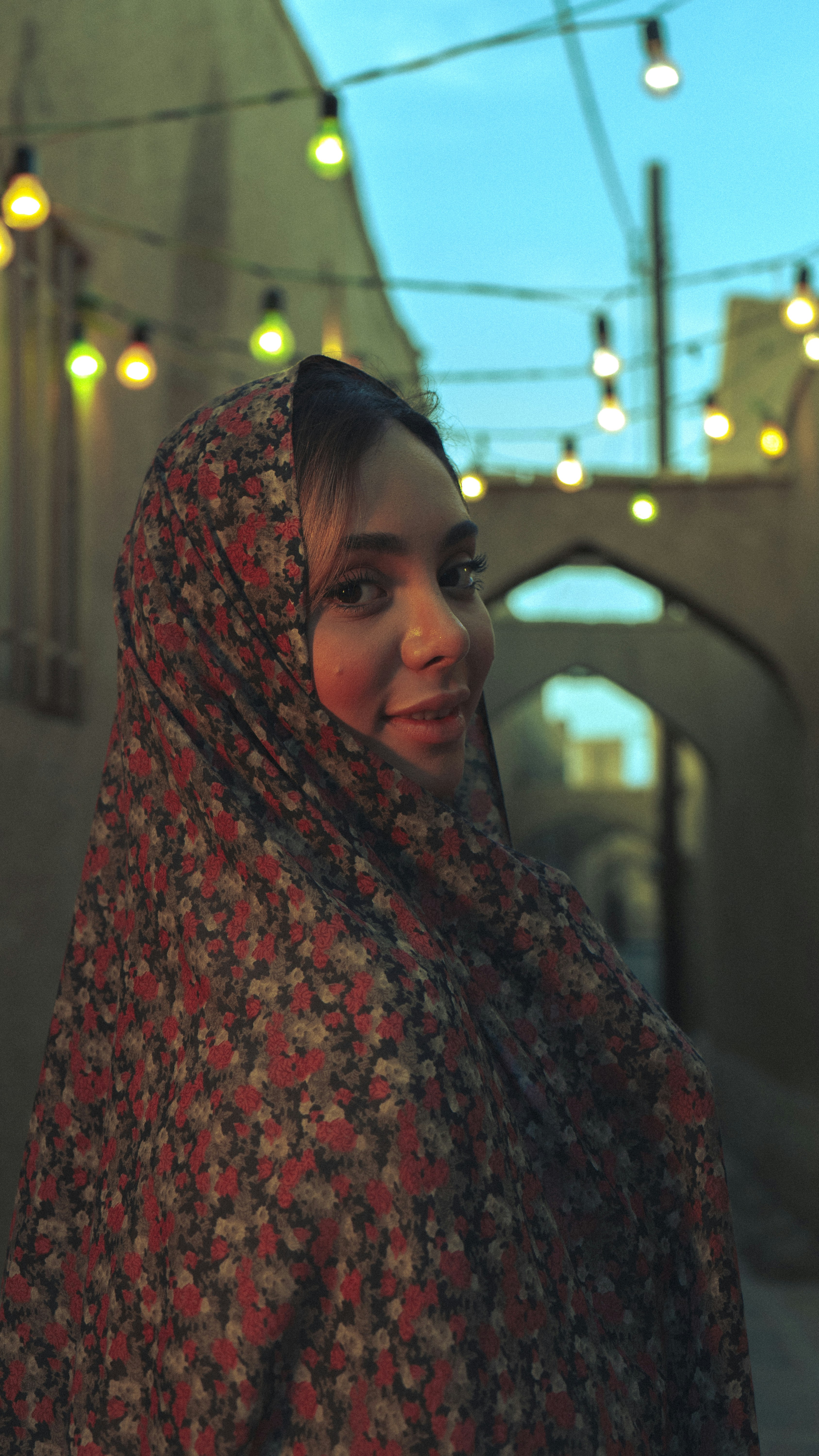 Woman in floral shawl stands in a narrow alley with colorful string lights overhead.