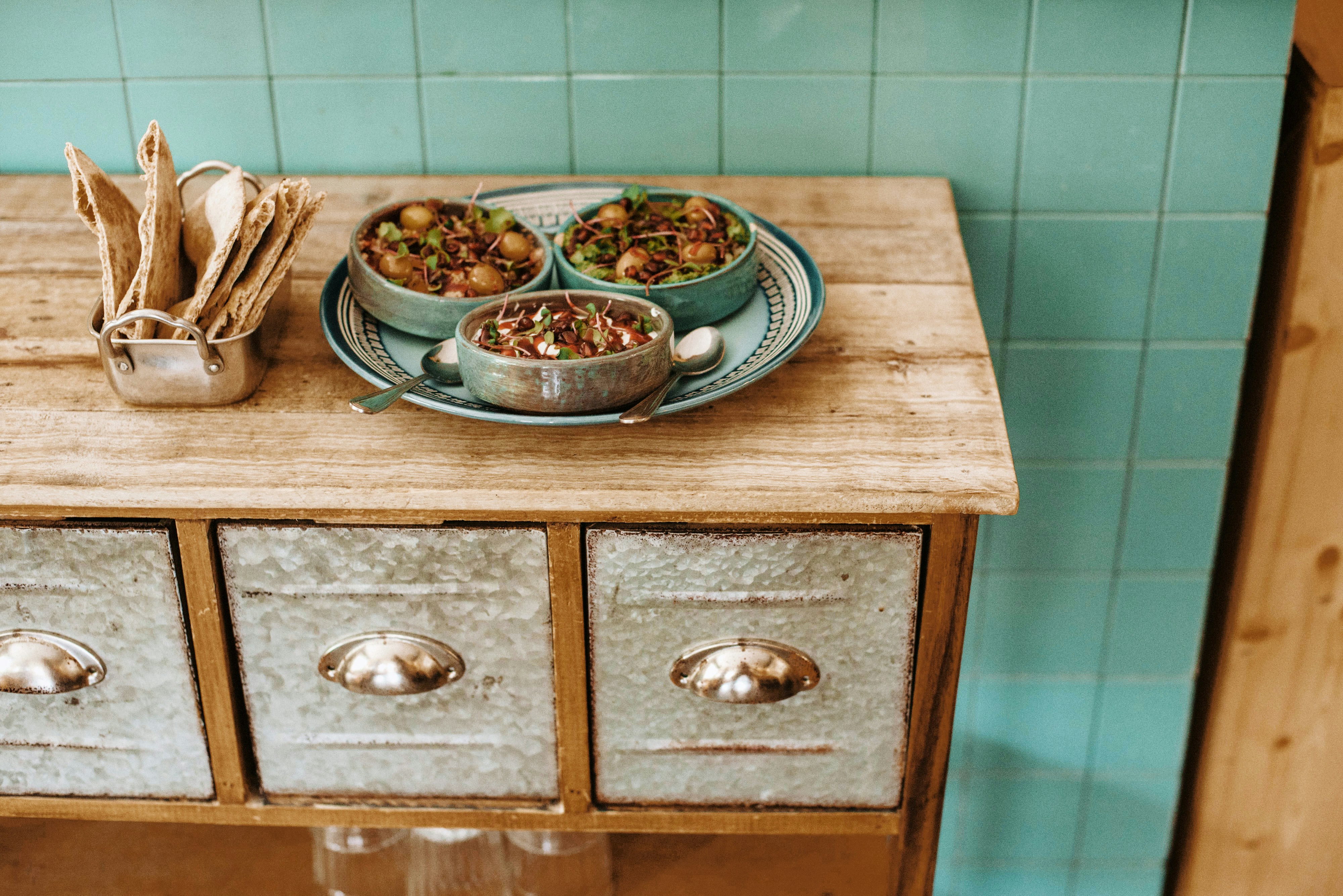 Rustic wooden sideboard with metal drawers topped with bowls of fresh salad against a teal tiled wall.