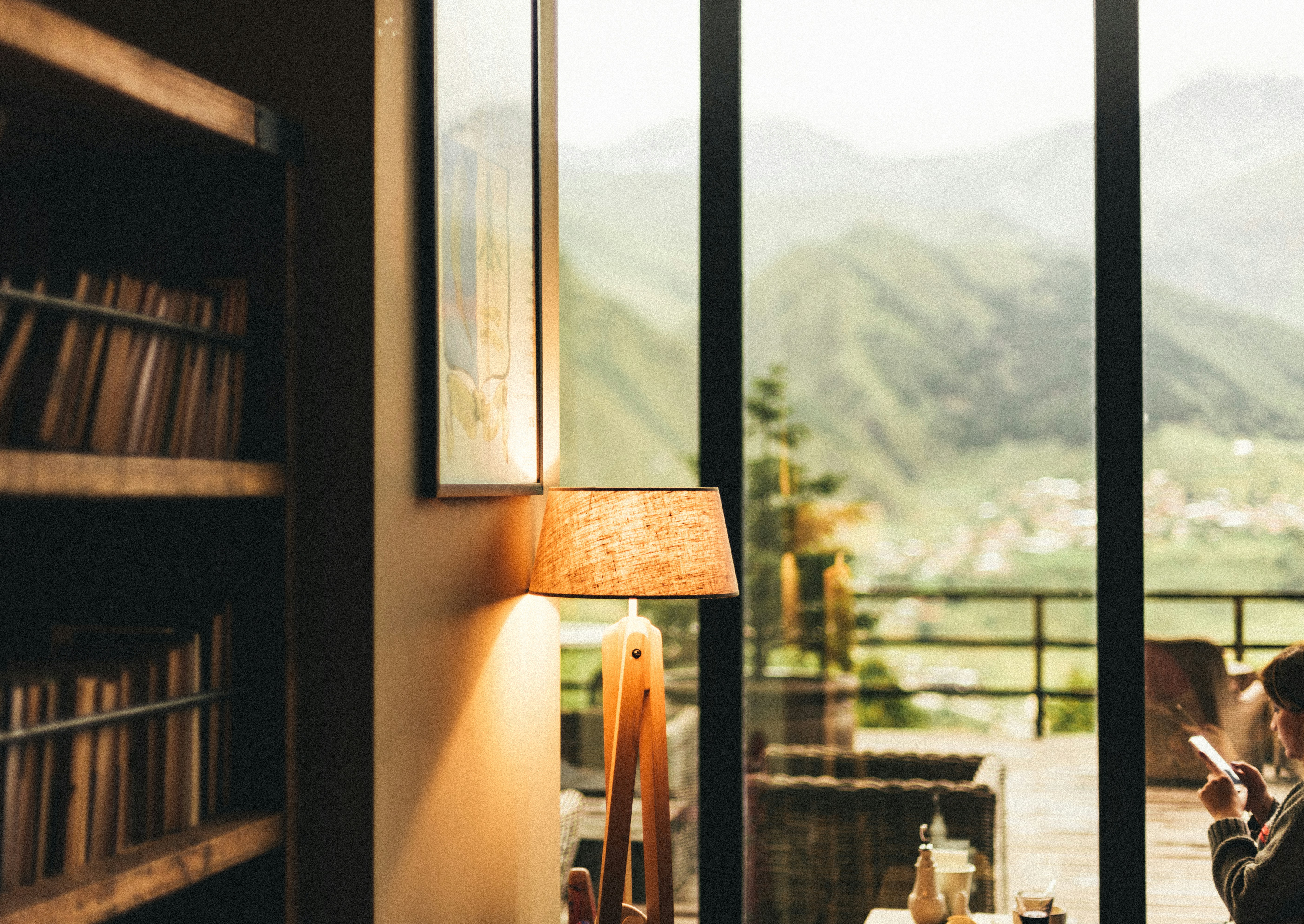 Cozy interior with bookshelves and a lamp, overlooking misty mountains through large windows.
