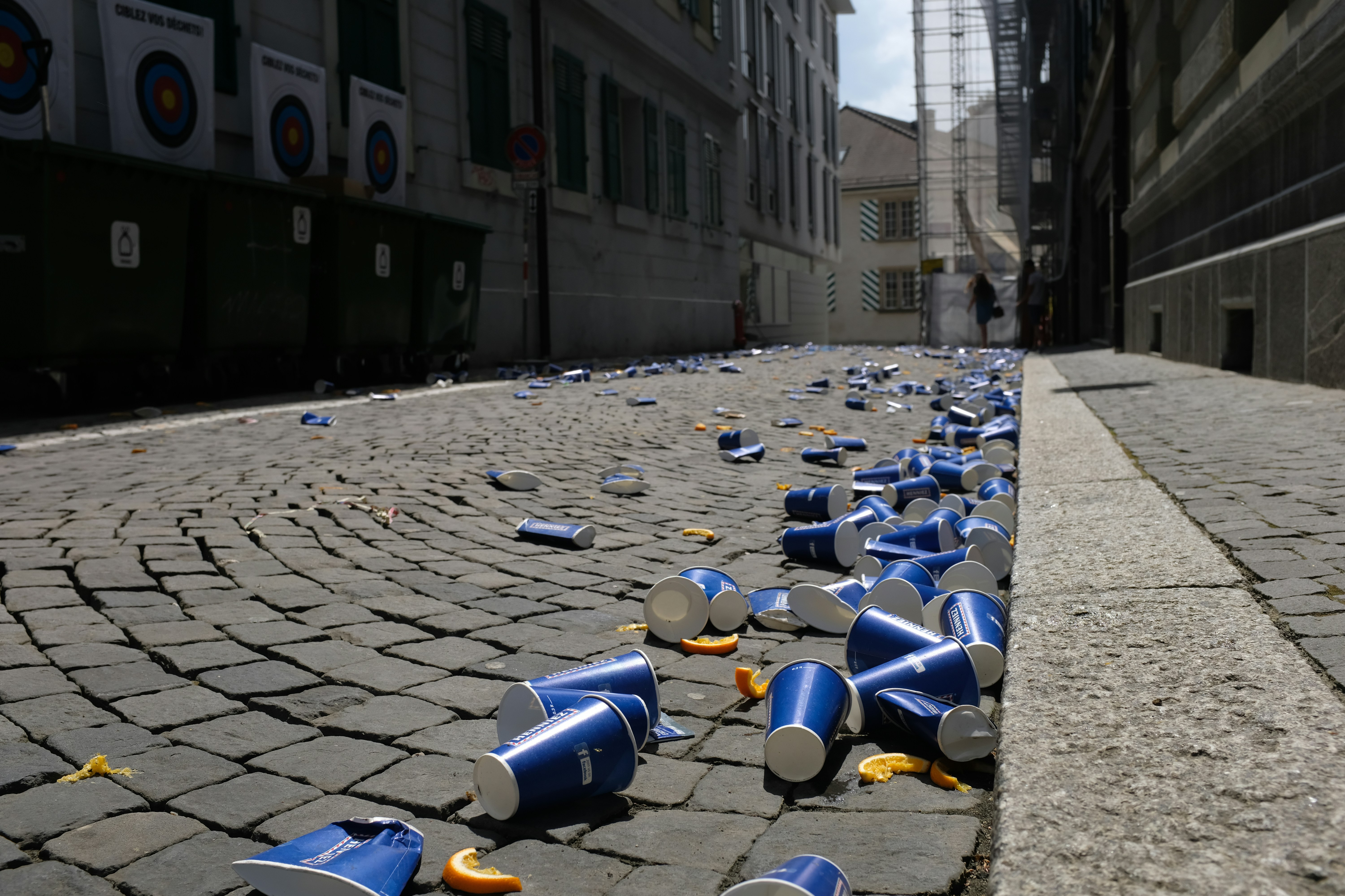 Scattered blue cups and remnants of a lively gathering litter a cobblestone alleyway, reflecting the aftermath of an event. Sunlight casts shadows, adding depth to the scene.