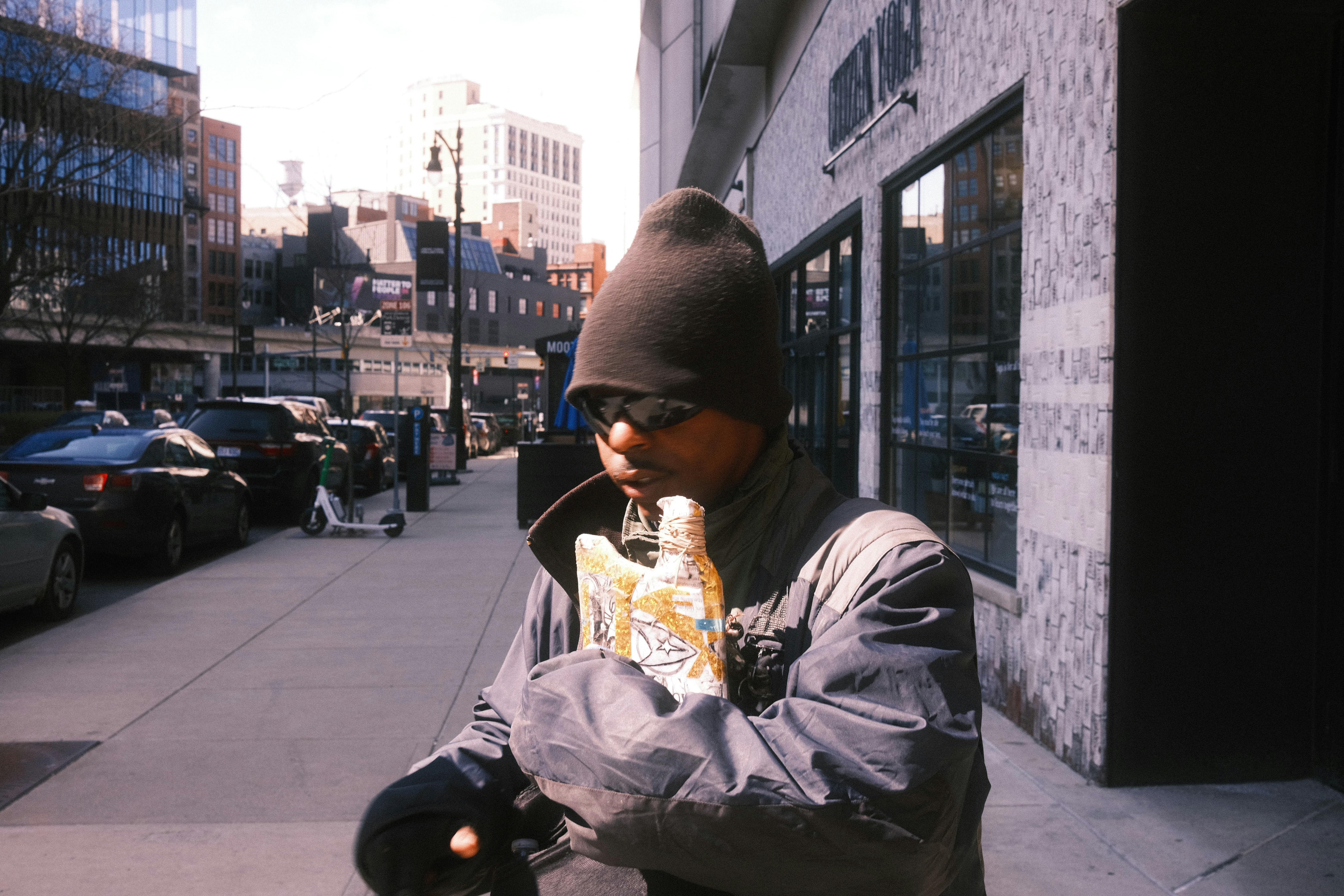 Person bundled in winter clothing walking on a city sidewalk, holding a snack package.