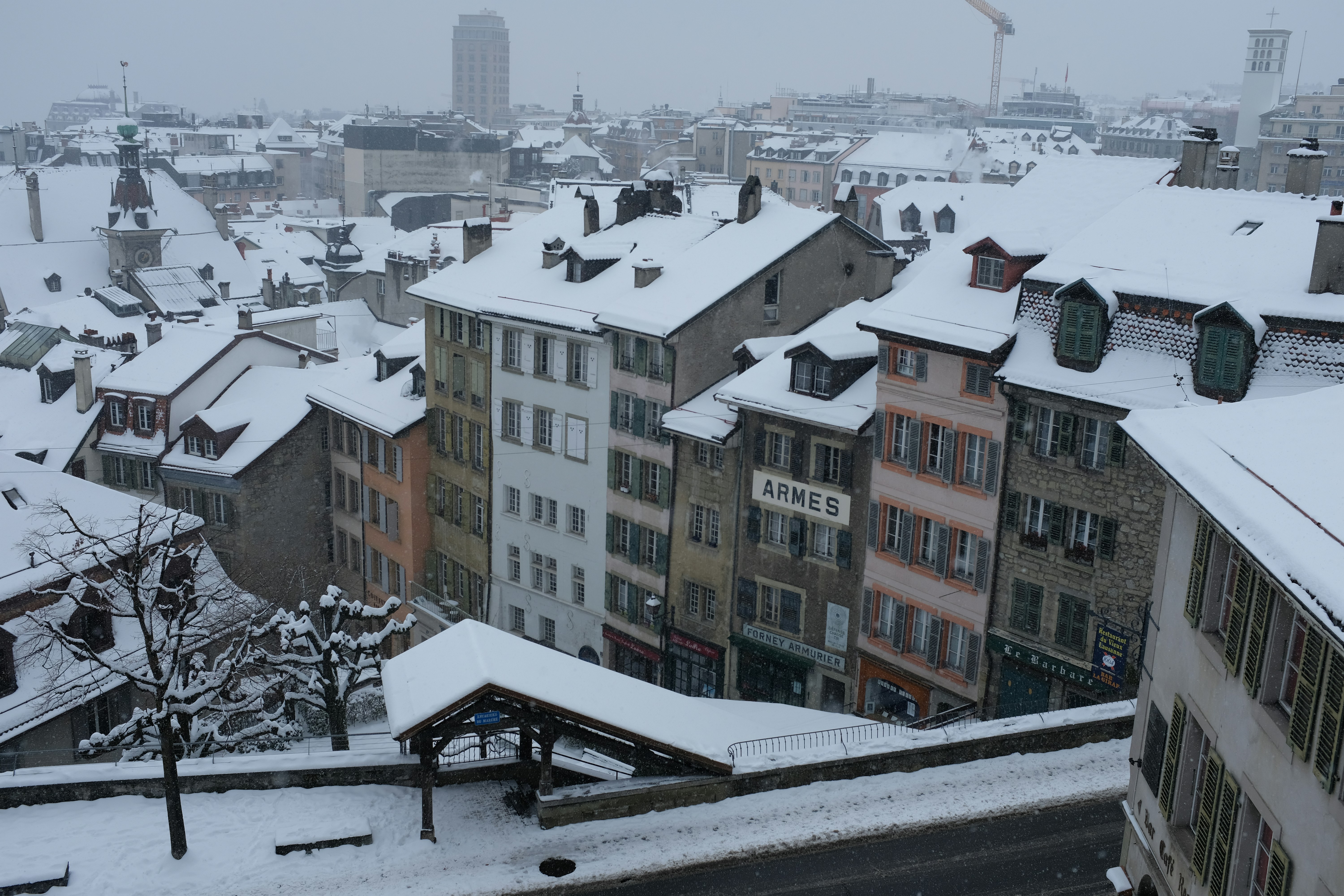 Snow-covered rooftops of charming buildings in a historic town, with a focus on the 'ARMES' sign. The scene captures the serene beauty of winter.