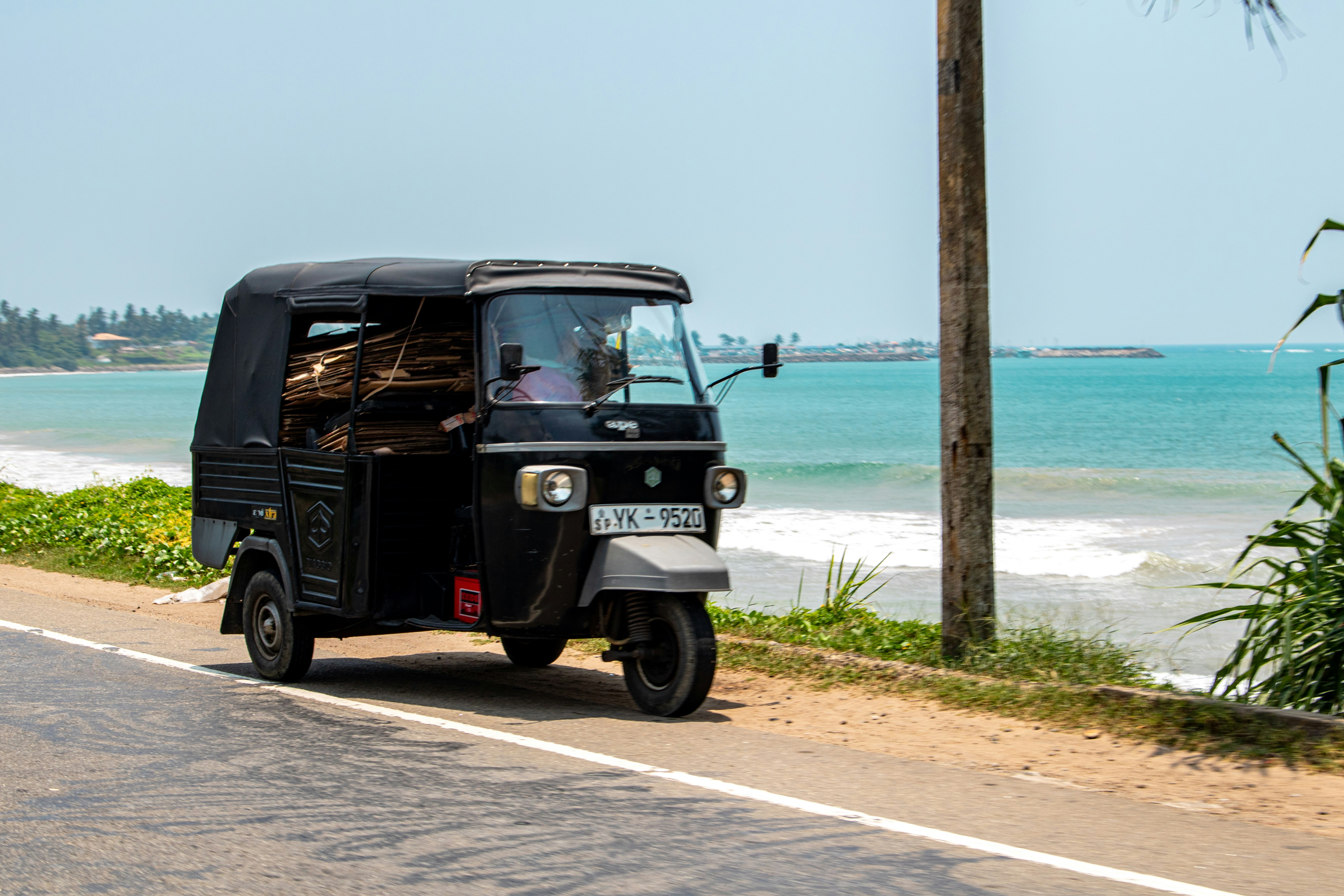 Black tuk-tuk parked on a coastal highway in Sri Lanka with ocean and greenery in the background.