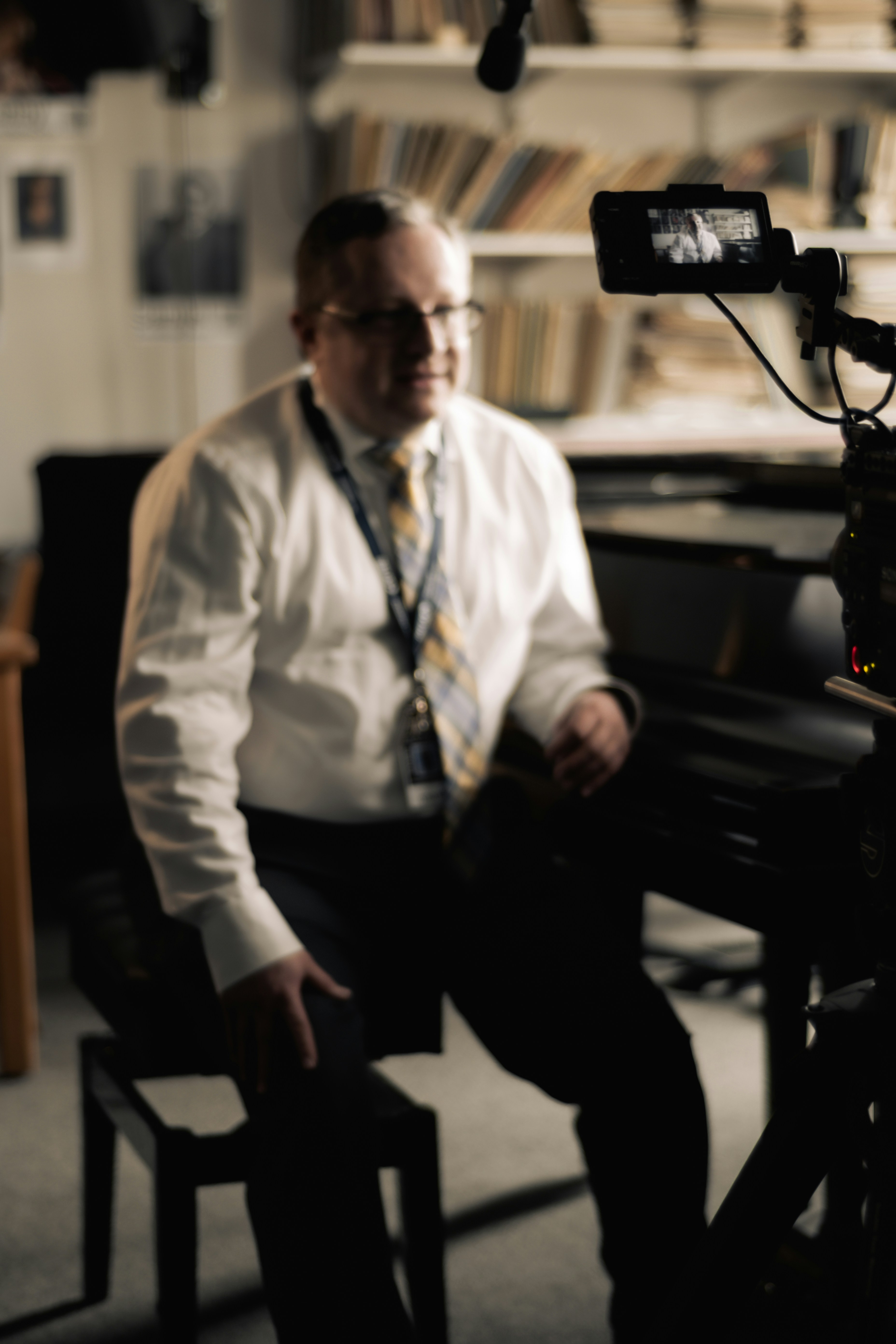 Man seated beside a piano in a dimly lit room, captured during an interview setup.