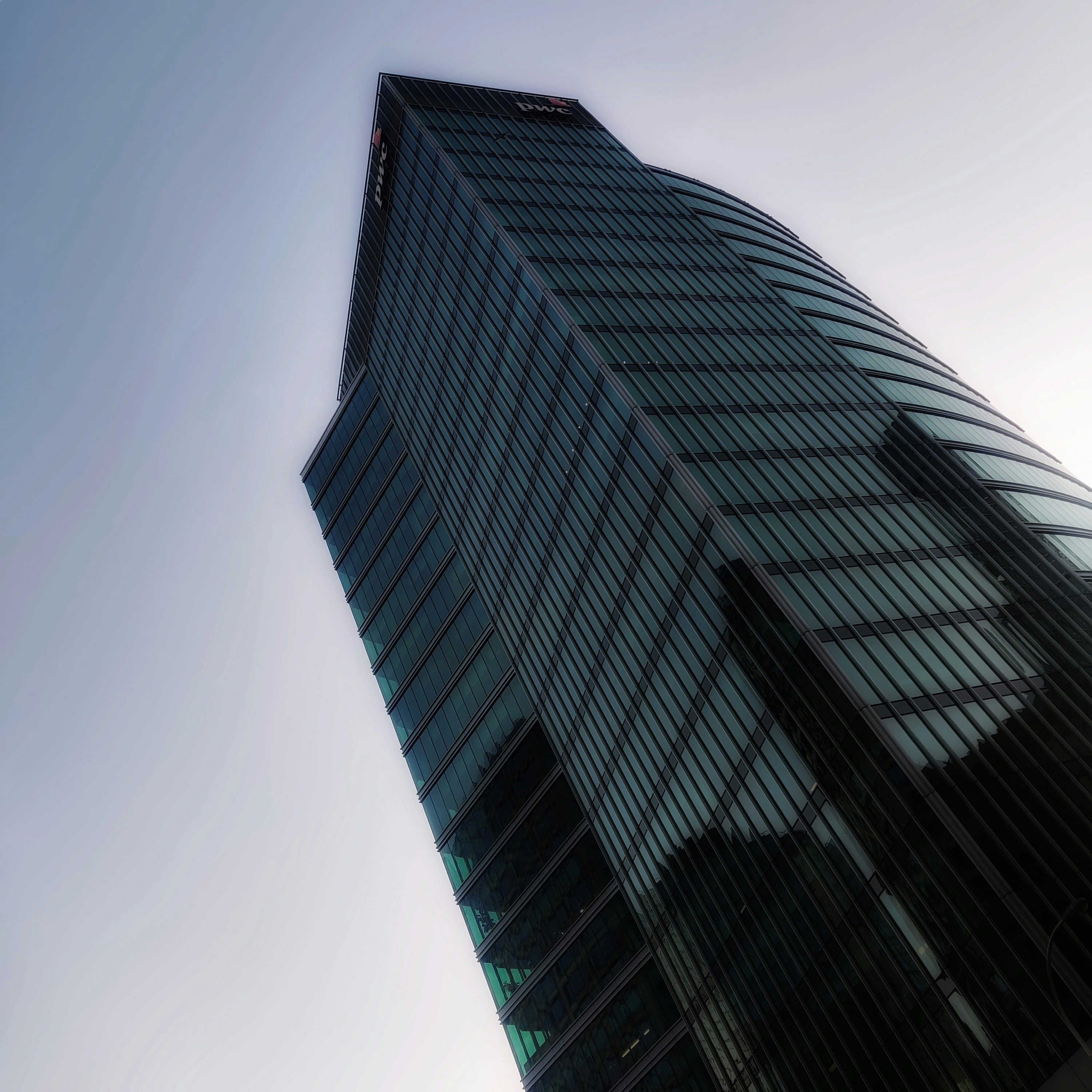 Low-angle photograph of a tall glass skyscraper with dark vertical lines and reflective panels against a pale sky.