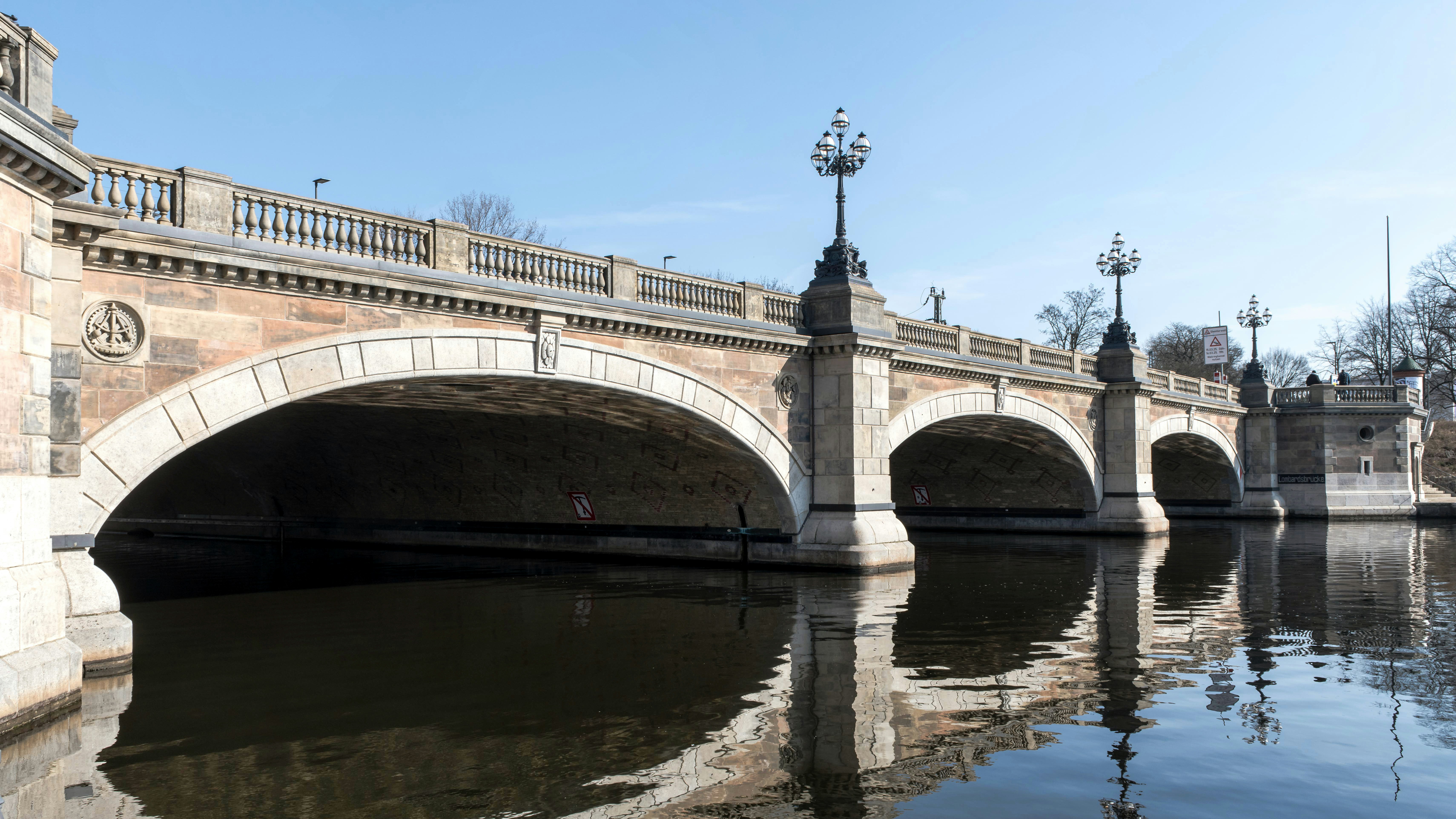 Stone bridge with ornate lampposts reflected in calm river under clear blue sky.