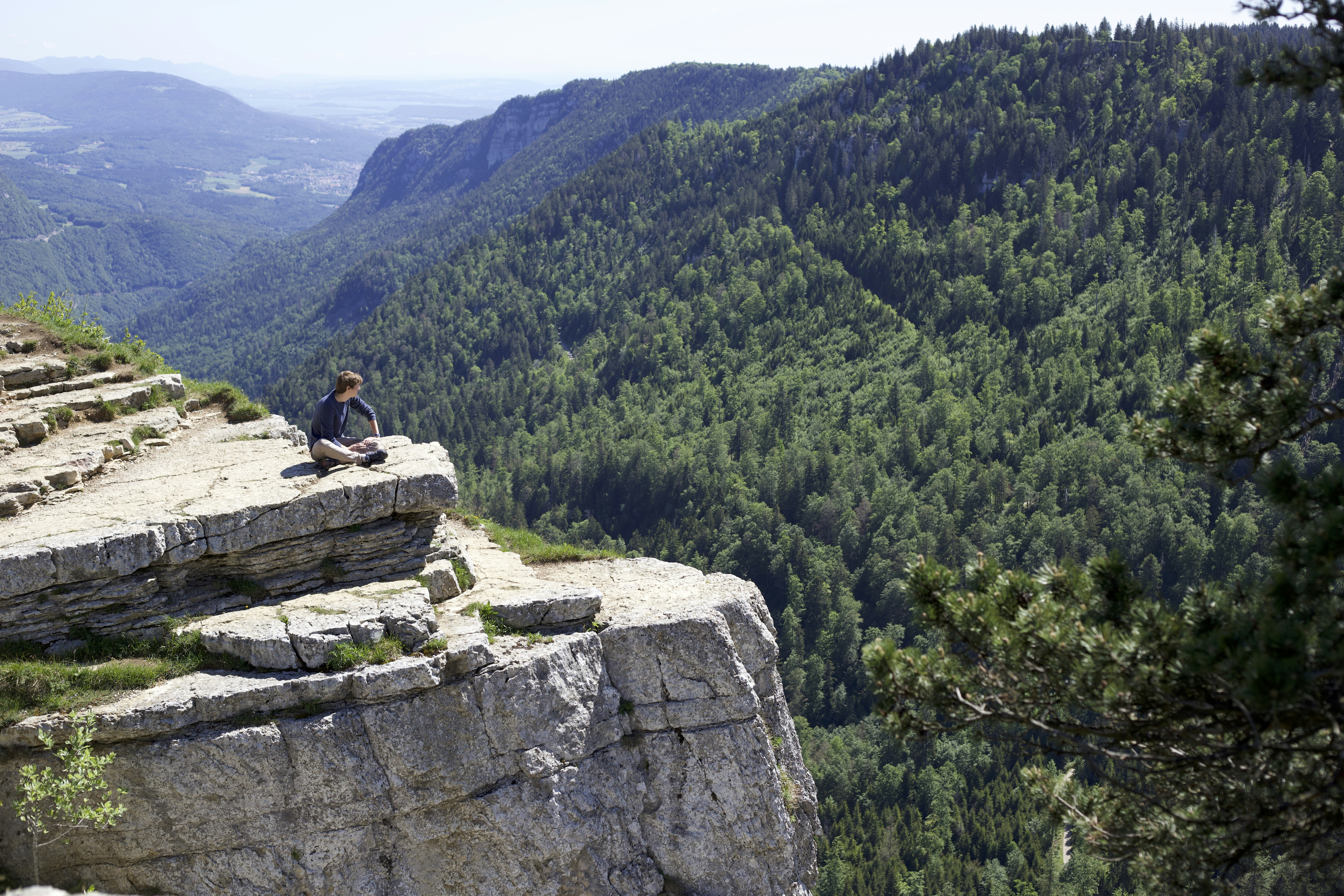 Person sitting on a rocky cliff overlooking a vast forested landscape under a clear sky.