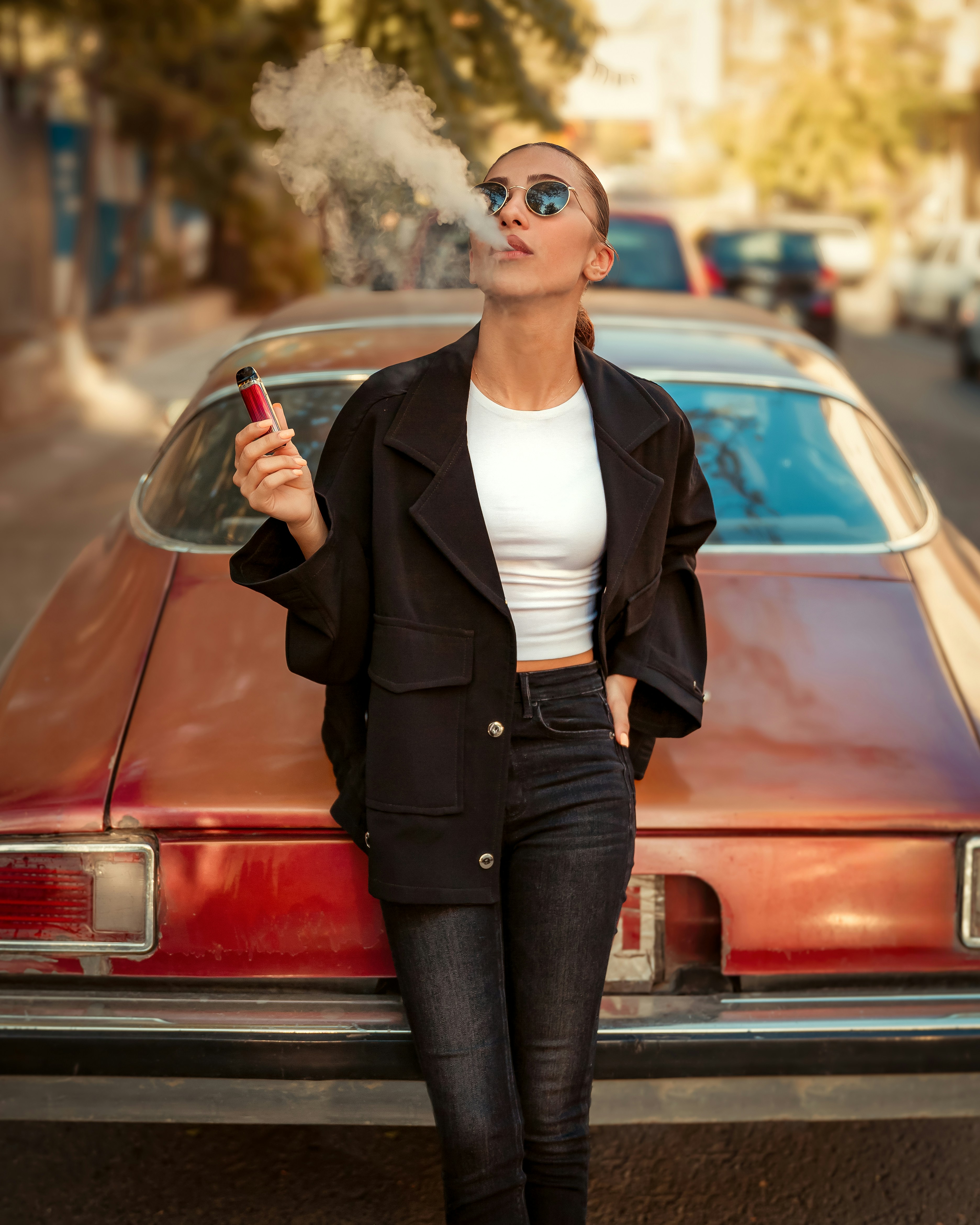 Woman leaning against a vintage car, exhaling vapor on a sunlit street.