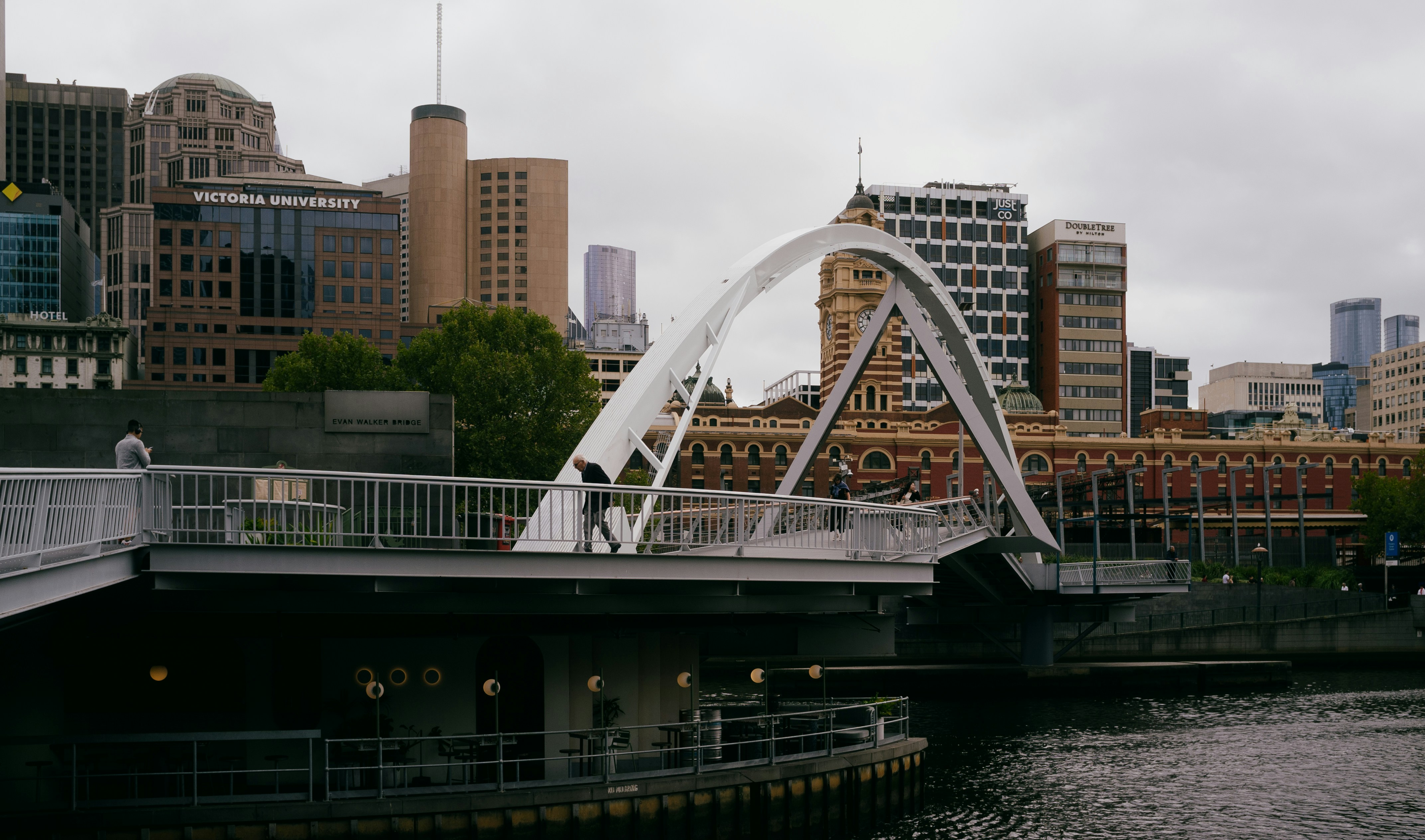 Modern arched bridge spanning a river with city buildings in the background on an overcast day.