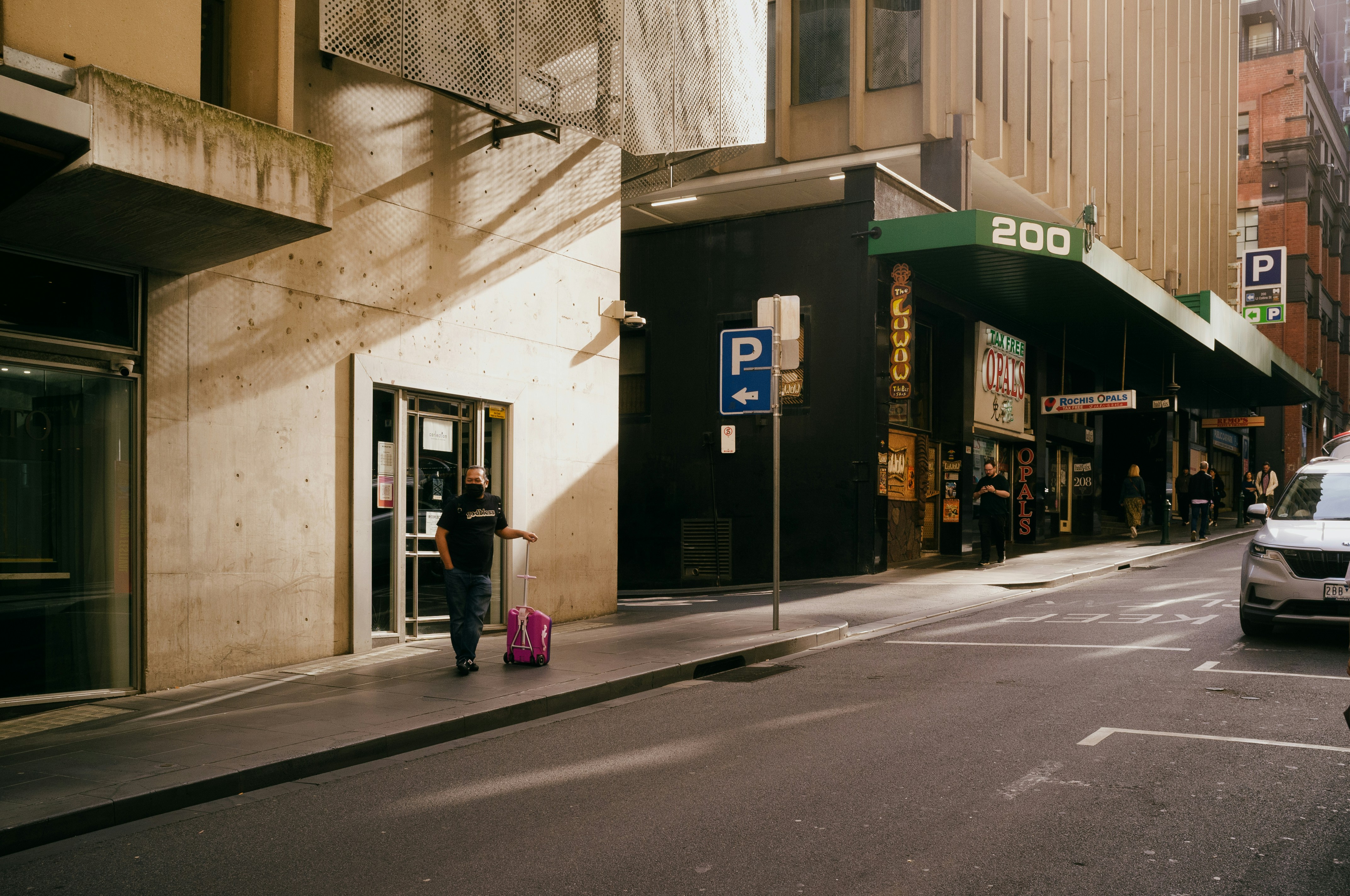 Person with a pink suitcase walking along a sunlit city street near parked cars.