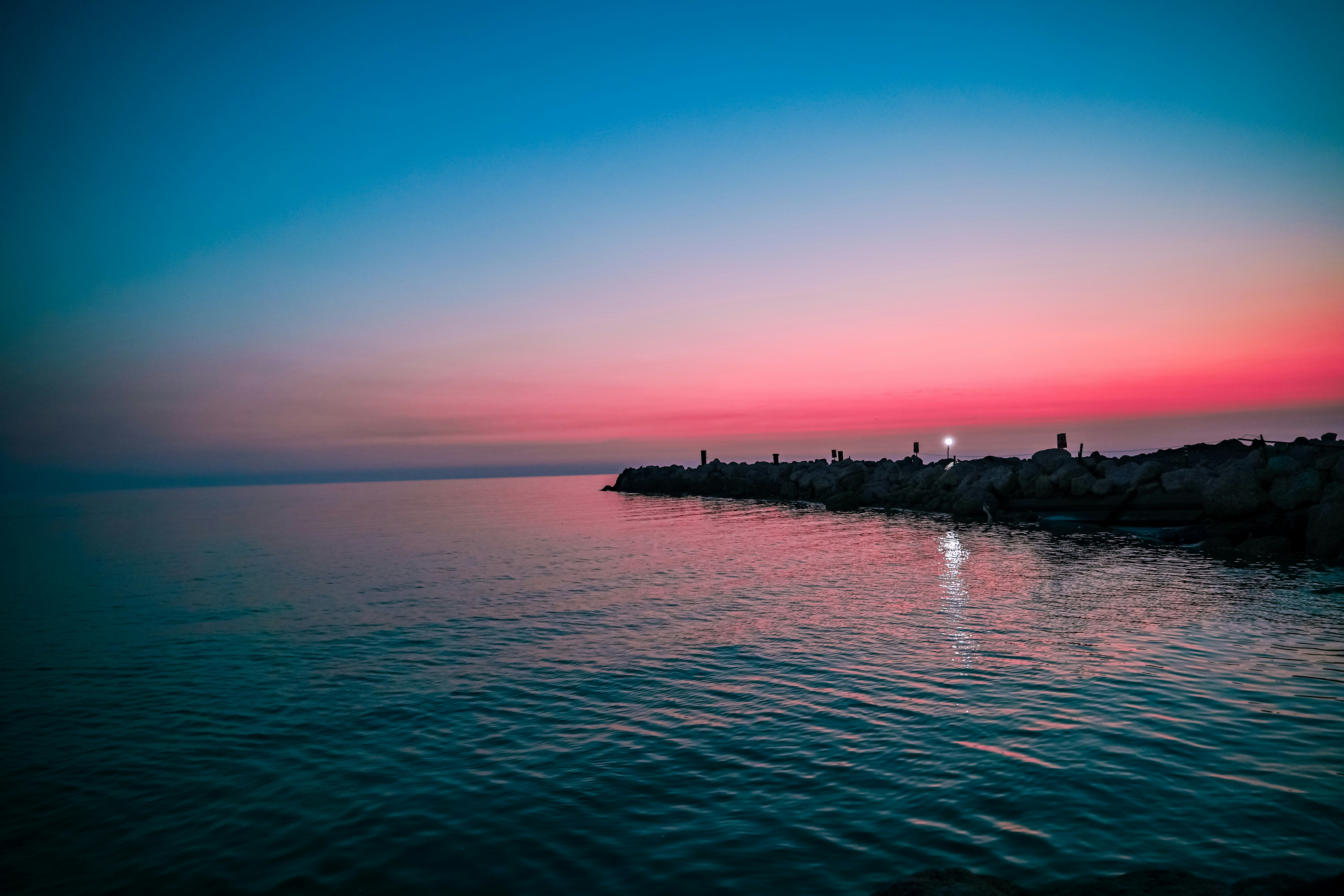 Vibrant sunset hues reflect on a calm sea beside a silhouetted pier with a distant lighthouse.