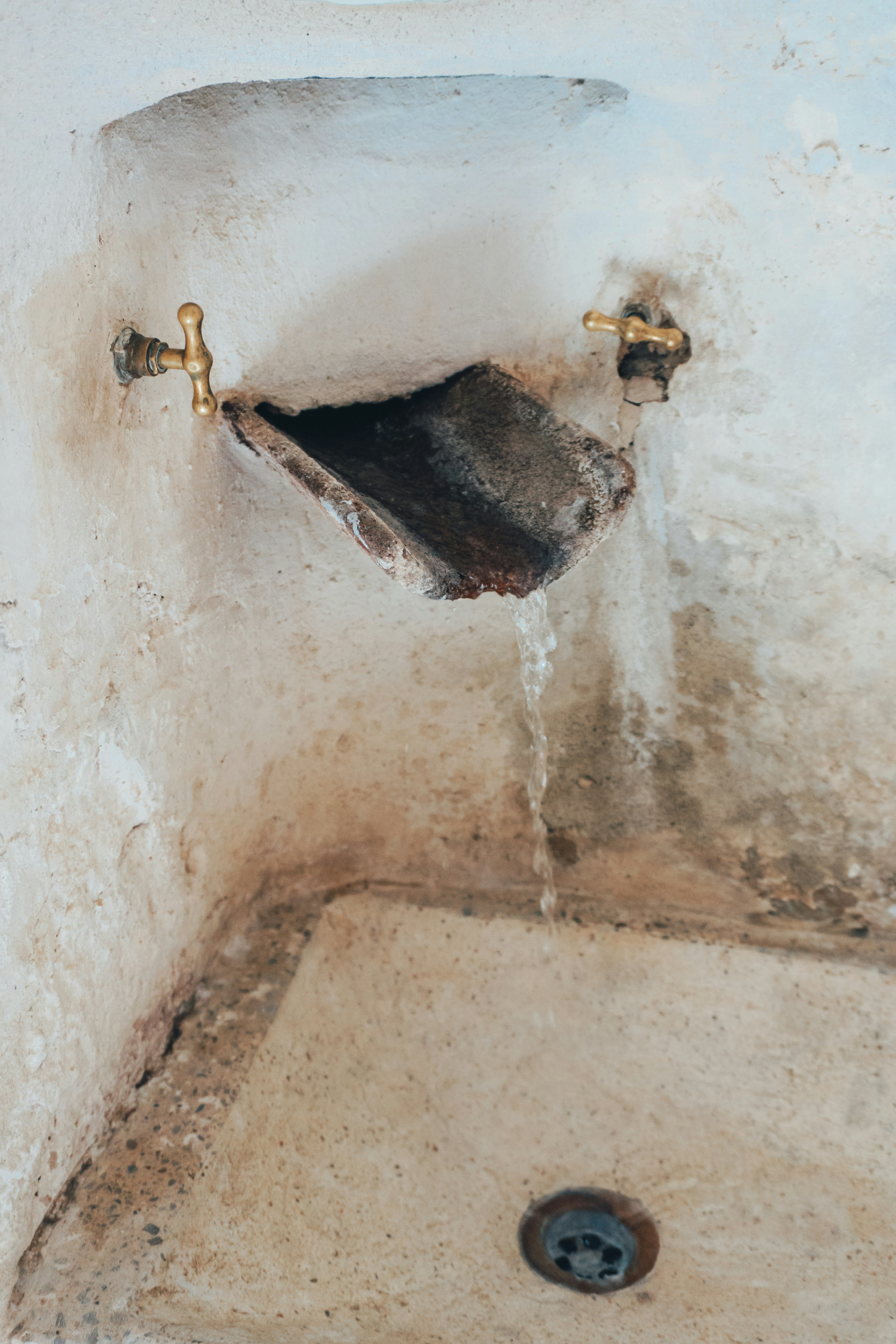 A weathered concrete sink with twin brass taps and a gaping wall hole leaking water, staining the basin. The scene highlights neglected utility and rugged, textured surfaces.