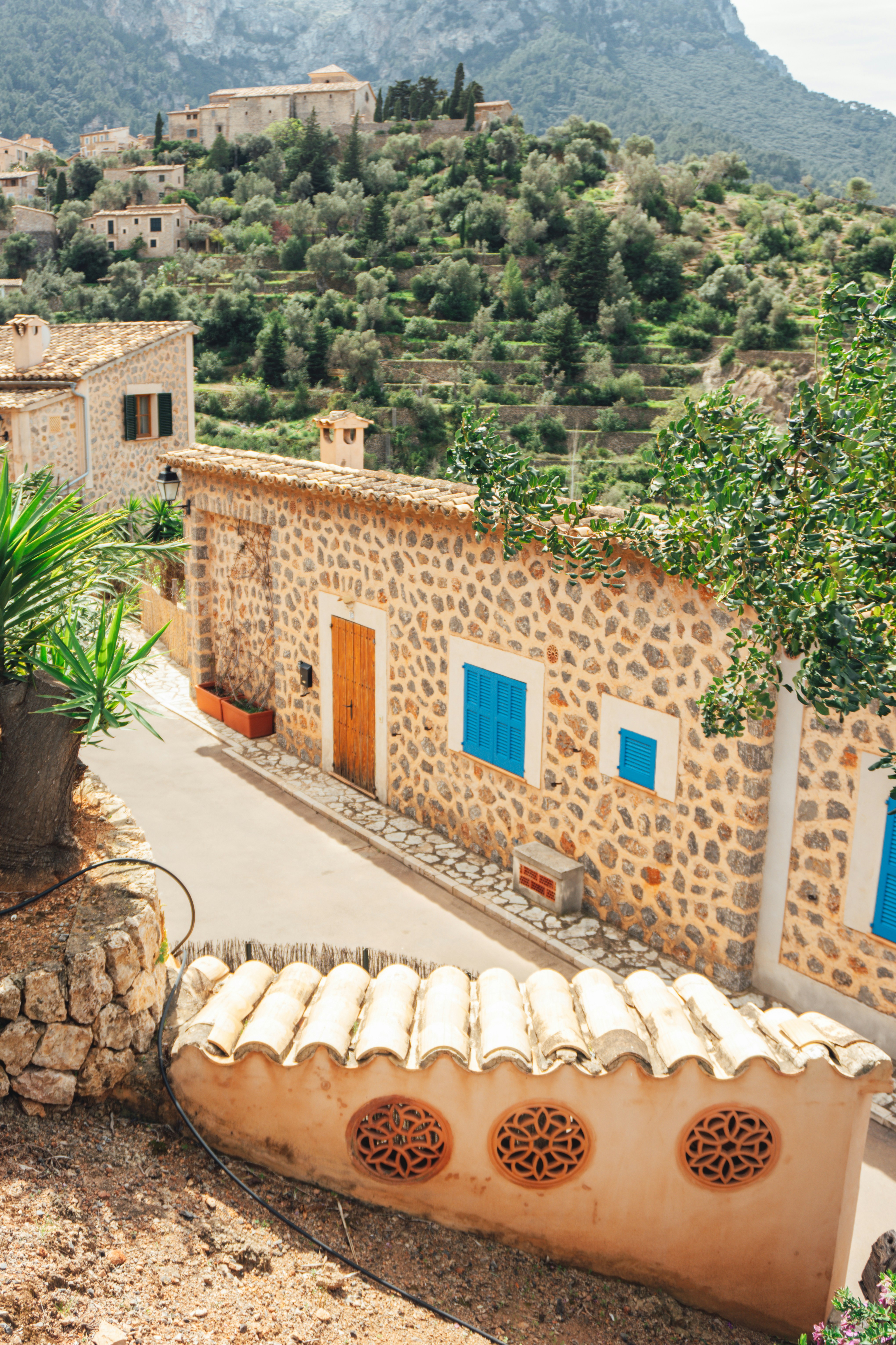 Charming stone buildings with blue shutters line a narrow road, set against lush terraced hills under a clear sky.