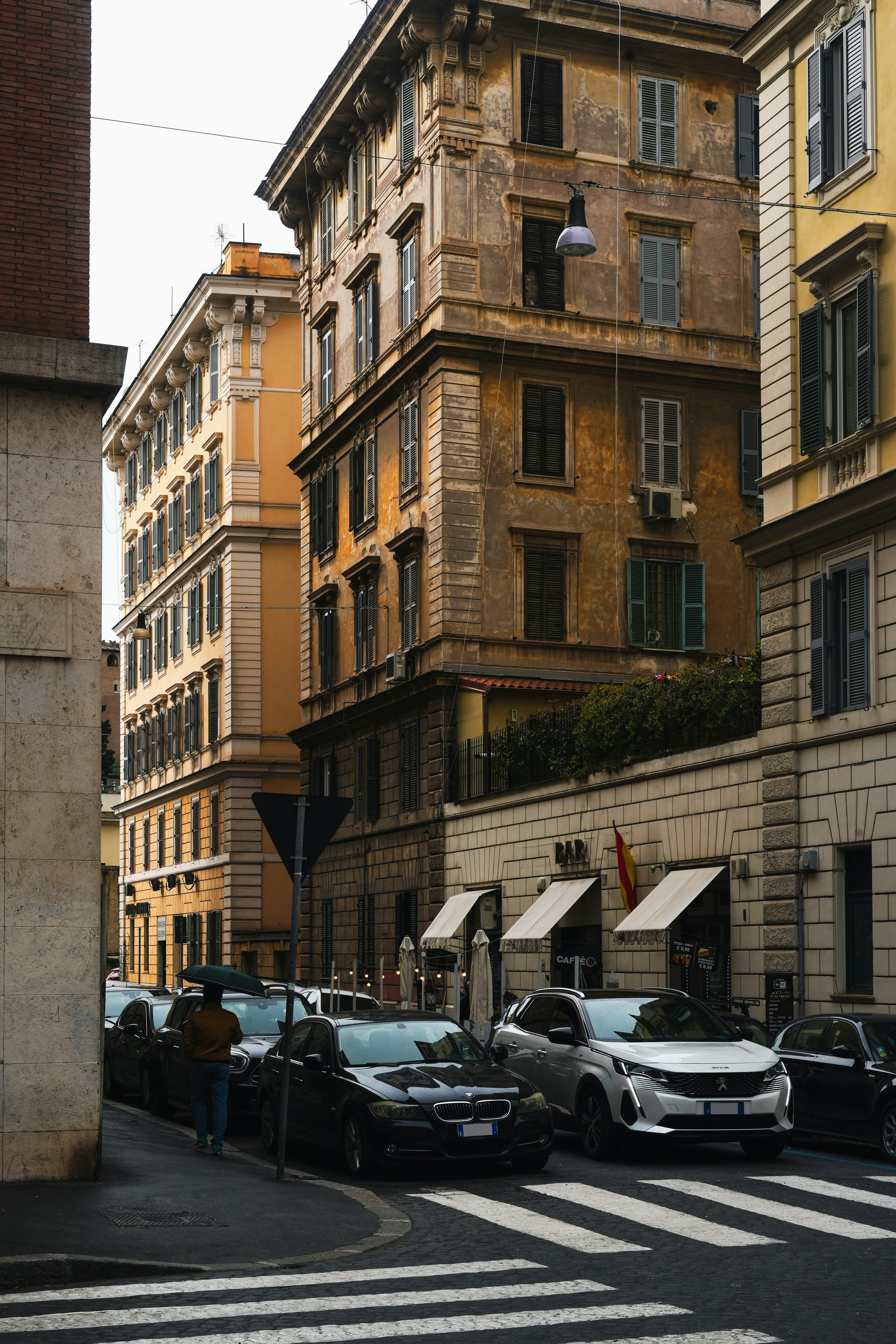 Historic buildings line a bustling street corner with parked cars and pedestrians on a cloudy day.