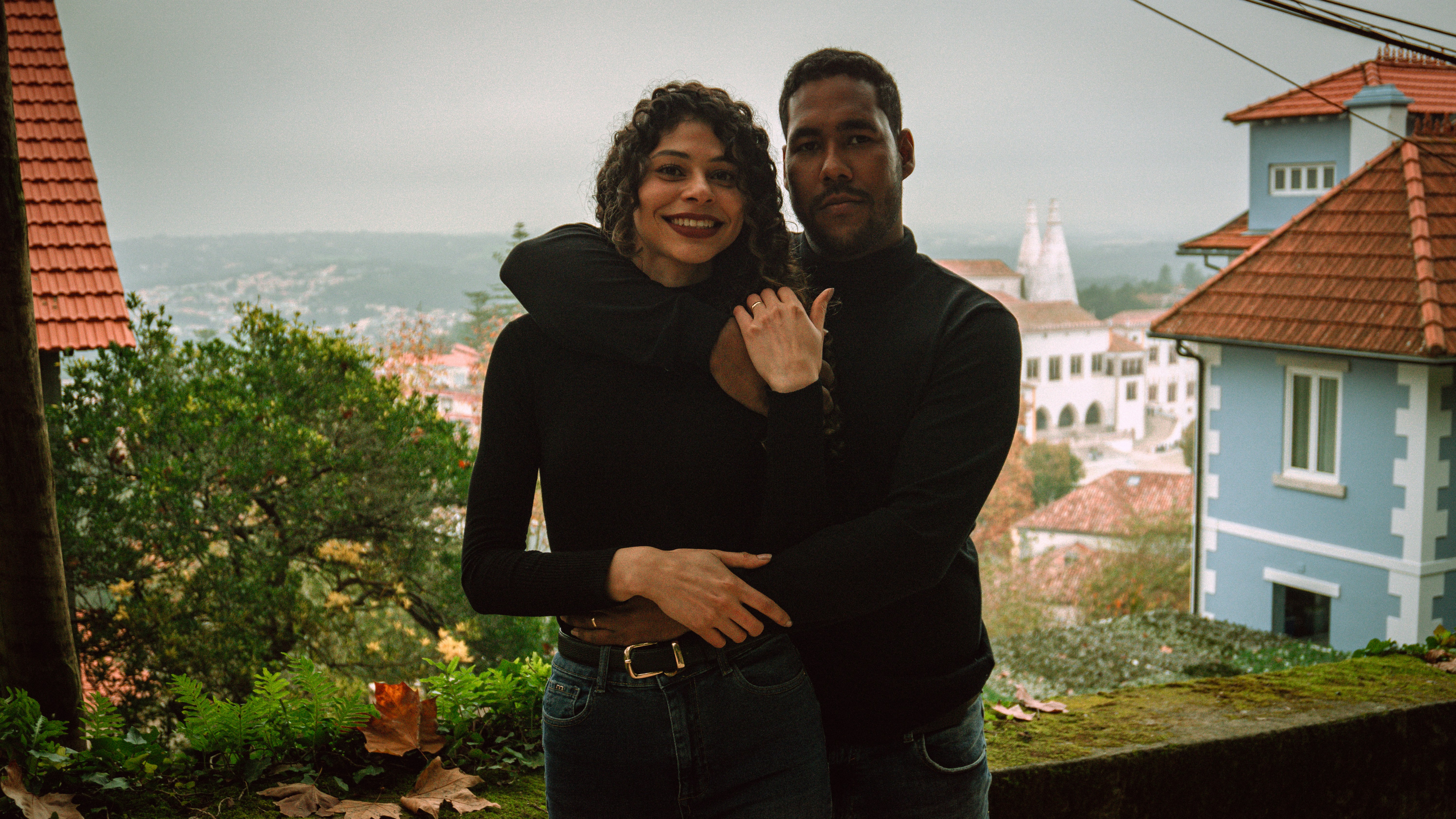 Couple embracing on a balcony with a scenic view of colorful rooftops and distant buildings under an overcast sky.