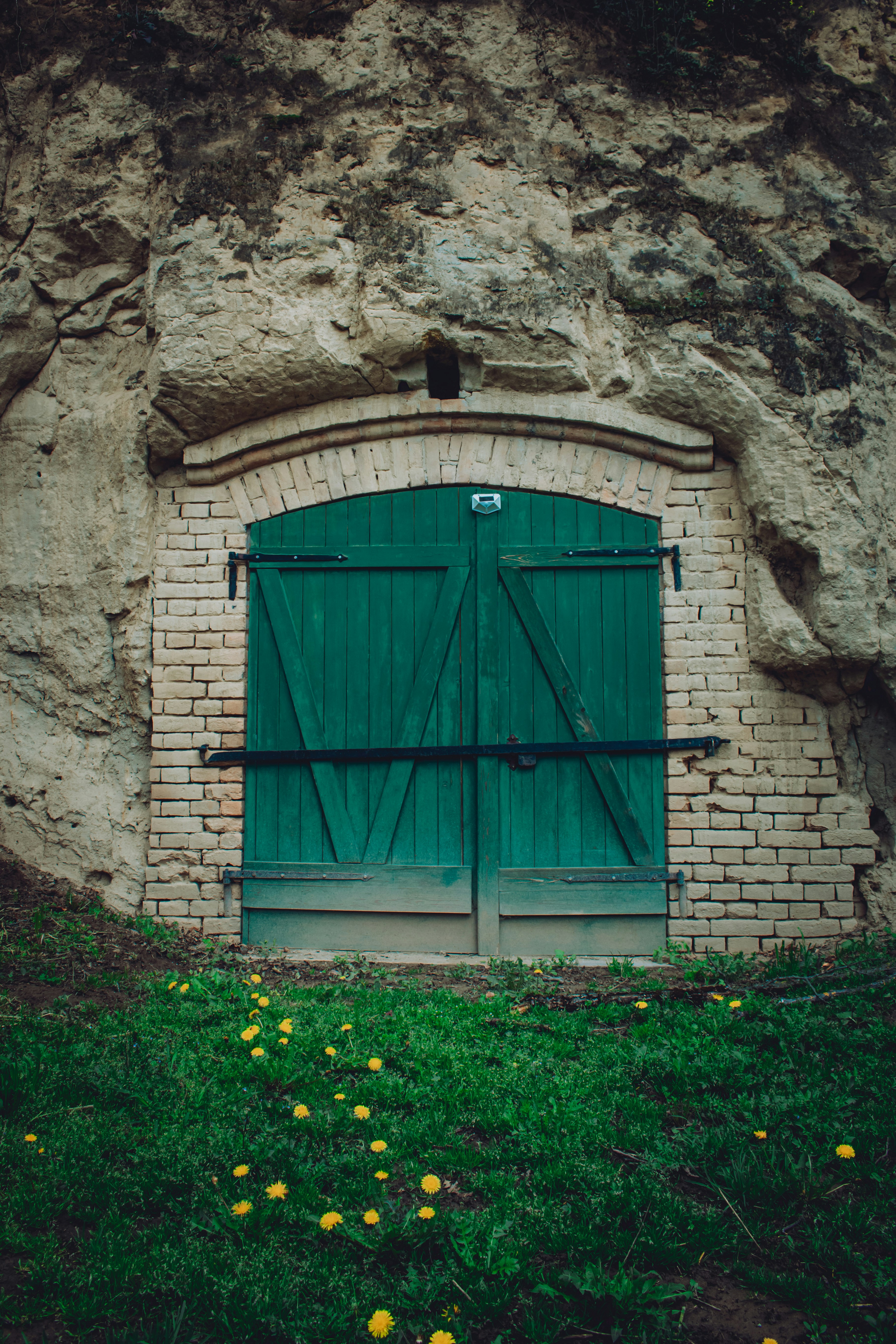 Green wooden doors set in a brick frame against a rocky hillside, with yellow flowers dotting the grass below.