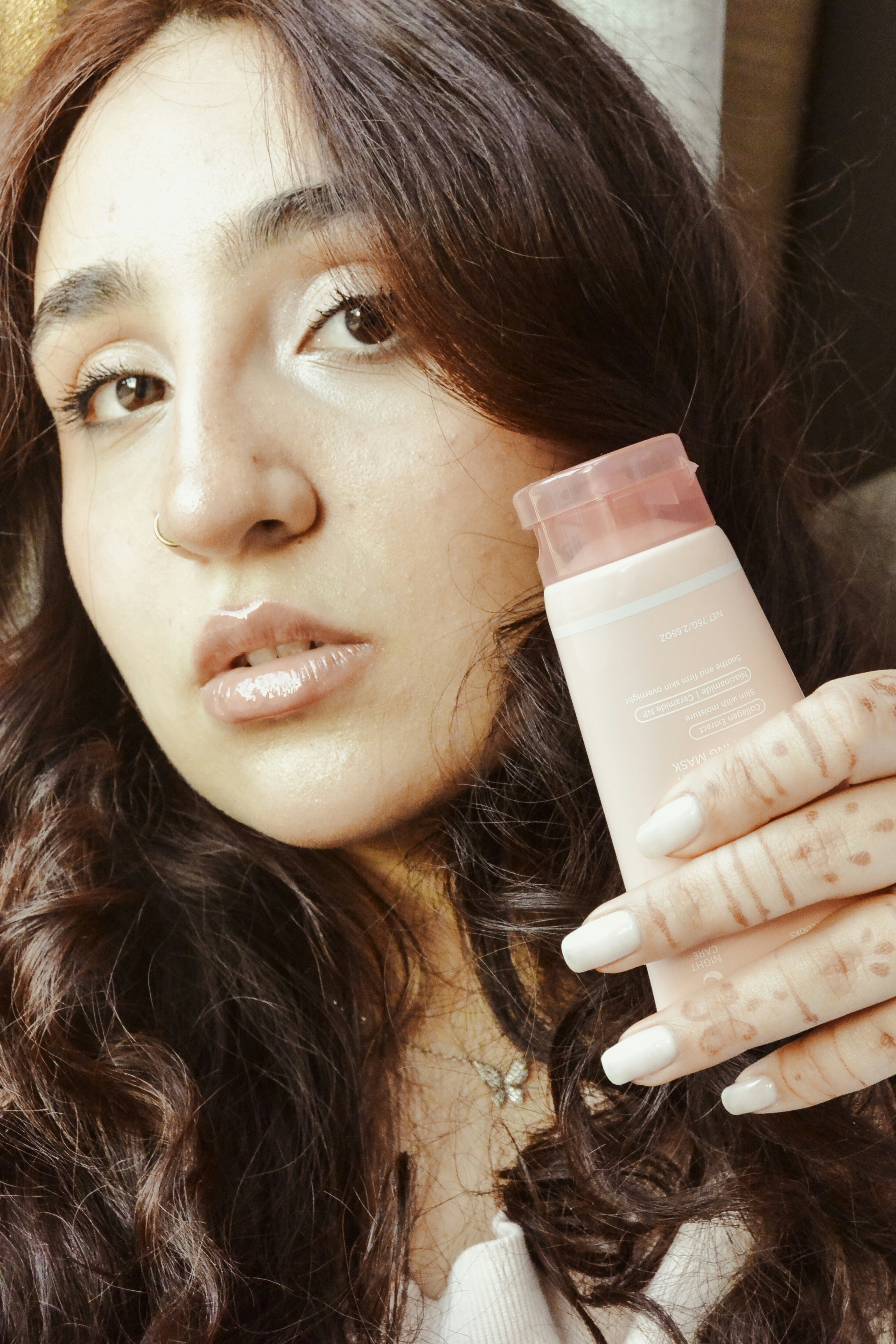 Woman with wavy brown hair holding a pink cosmetic bottle, lit by soft natural light.