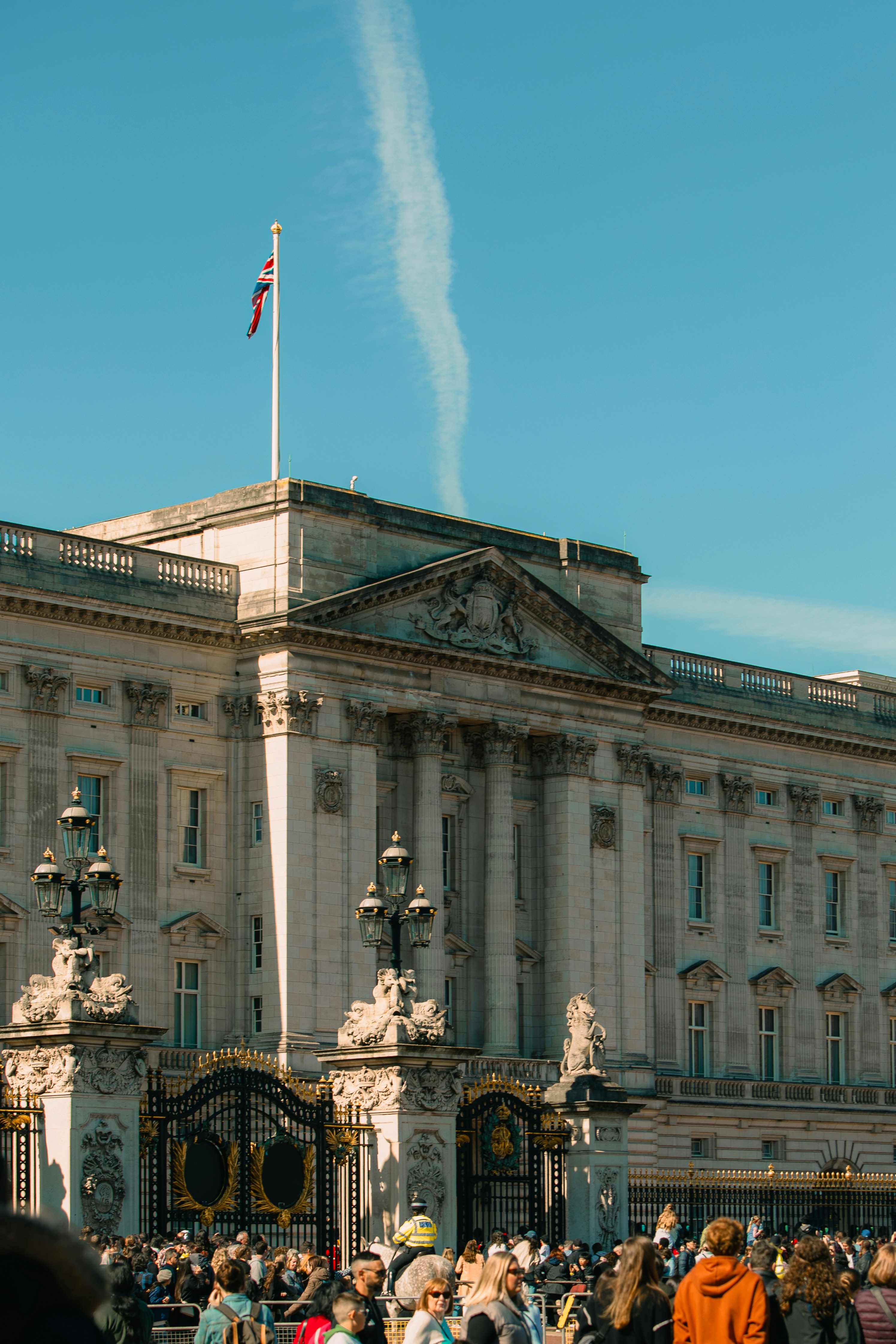 Large crowd outside Buckingham Palace on a sunny day with a jet stream in the clear sky.