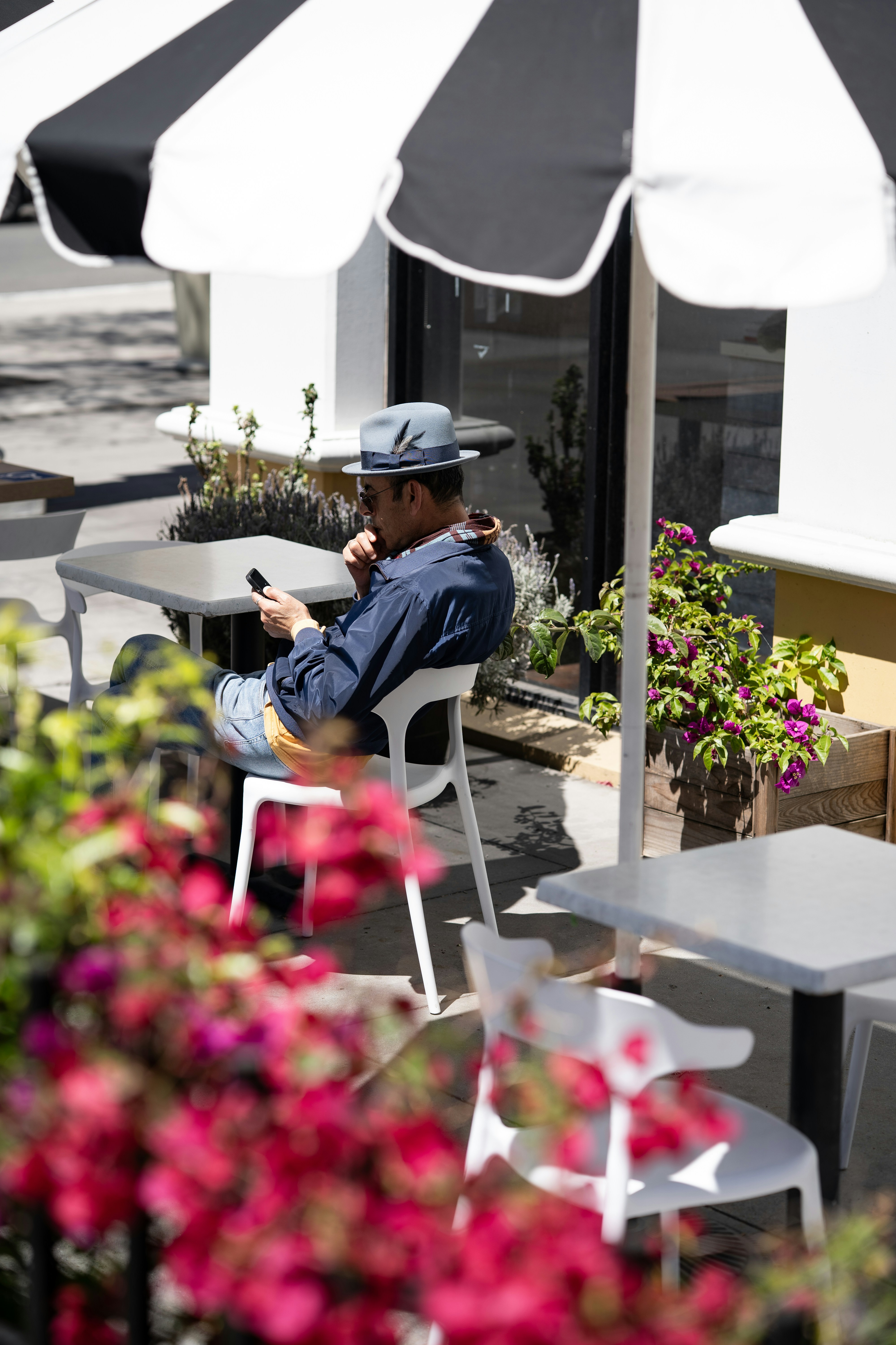 Man in a hat seated at an outdoor café under a striped umbrella, surrounded by blooming flowers.