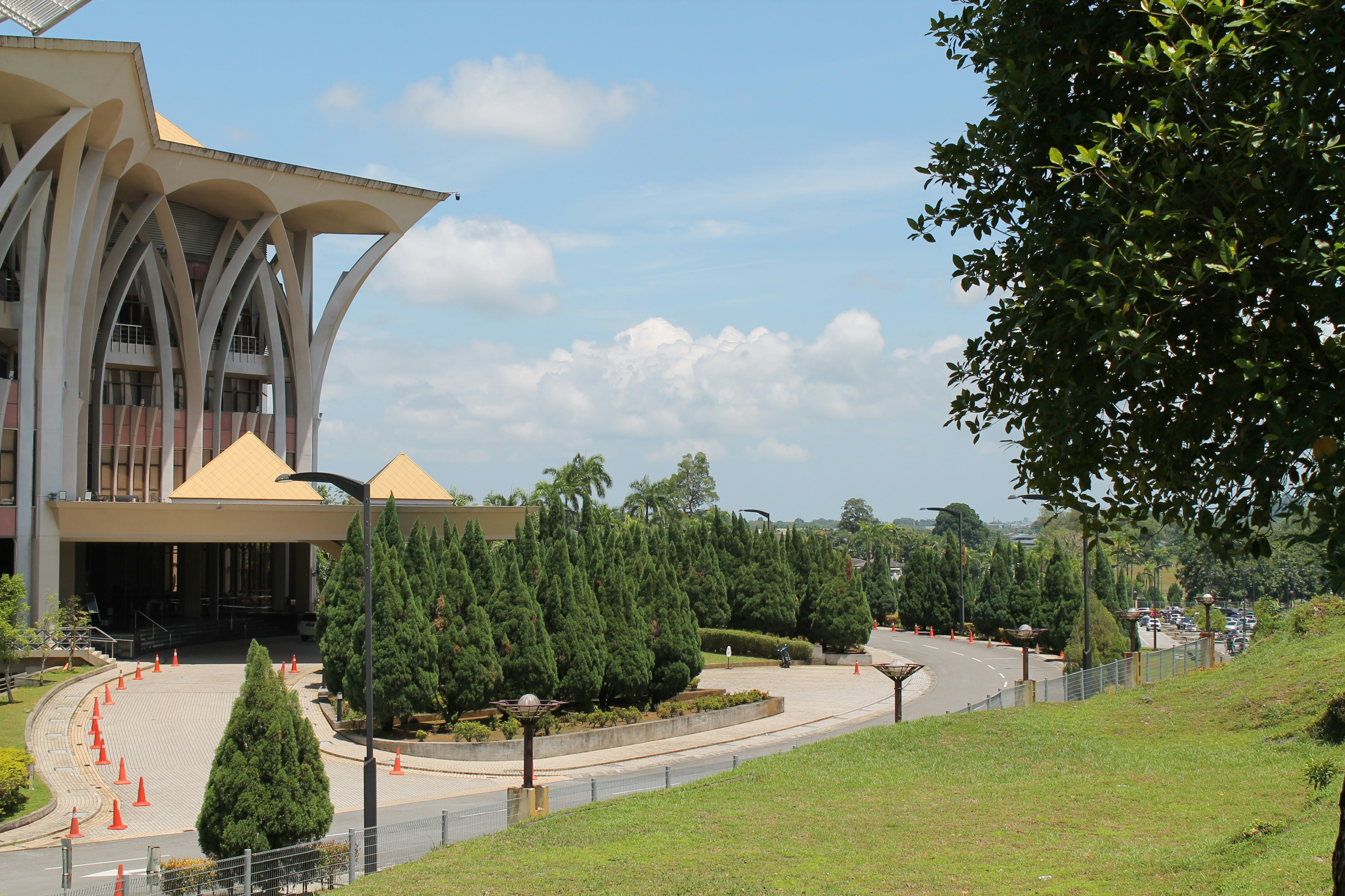 Contemporary building with arches beside a landscaped garden and winding road under a blue sky.