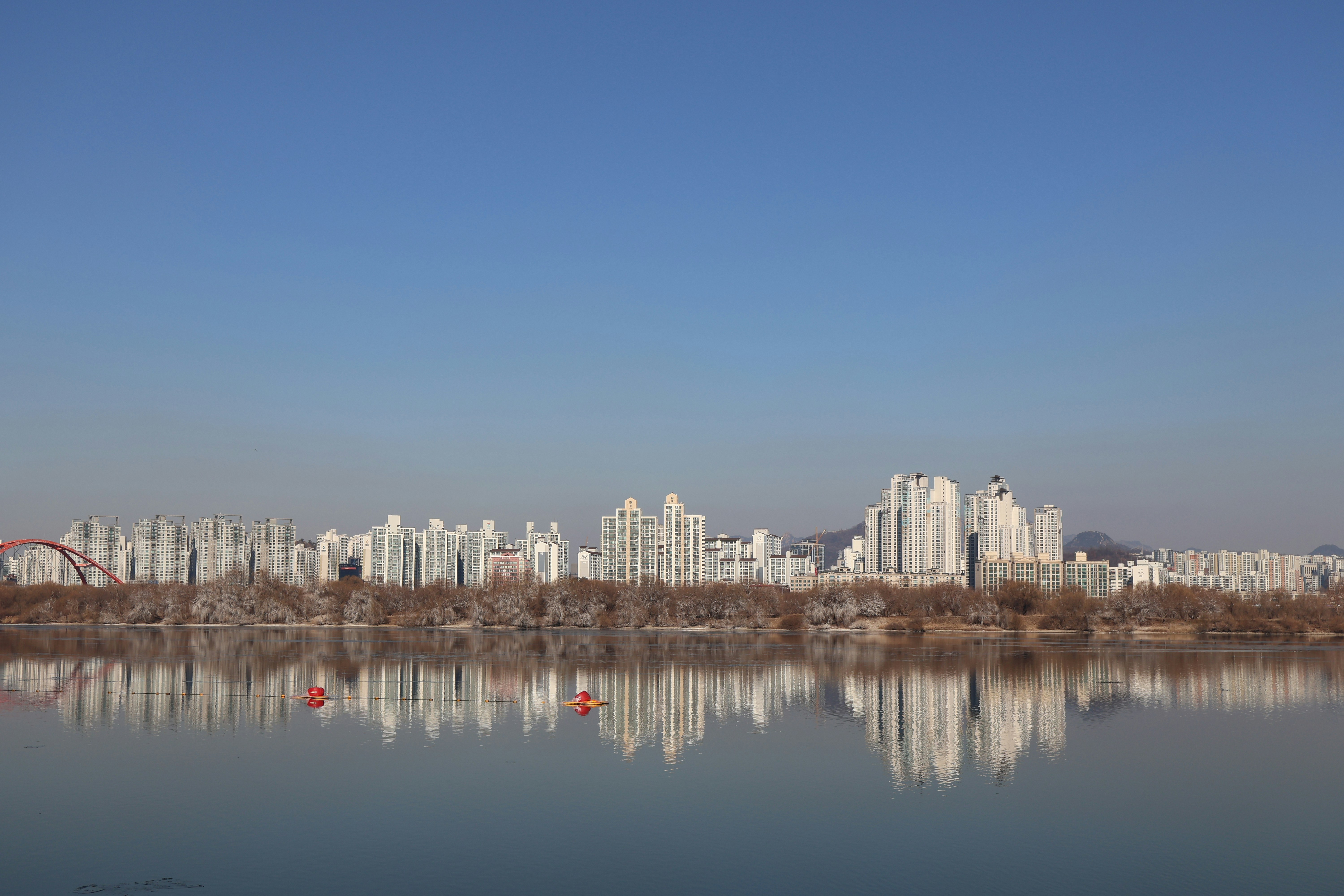 Modern city skyline mirrored in a calm river under a clear blue sky.