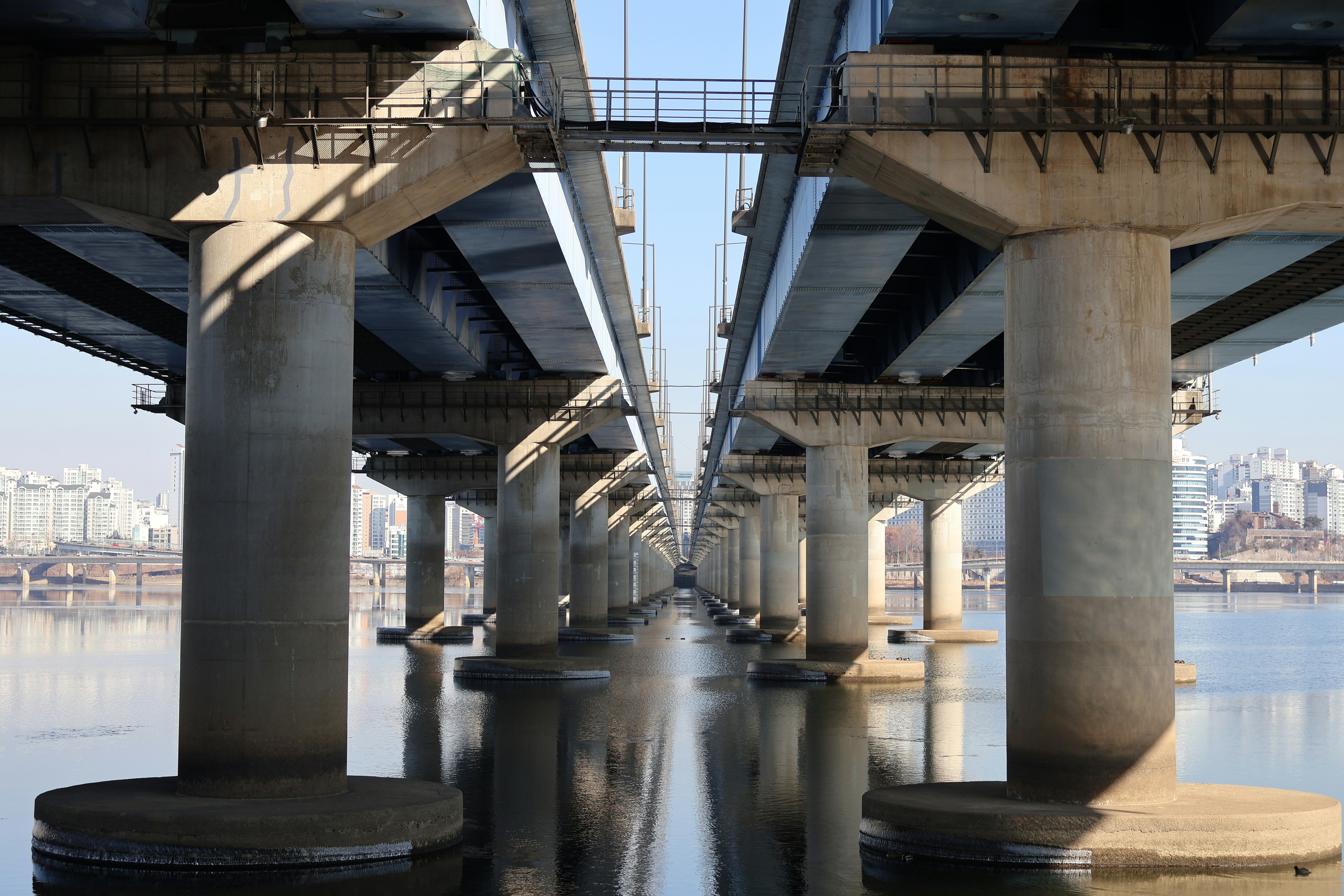 Underneath two parallel bridges spanning a calm river, showcasing architectural symmetry and reflections.