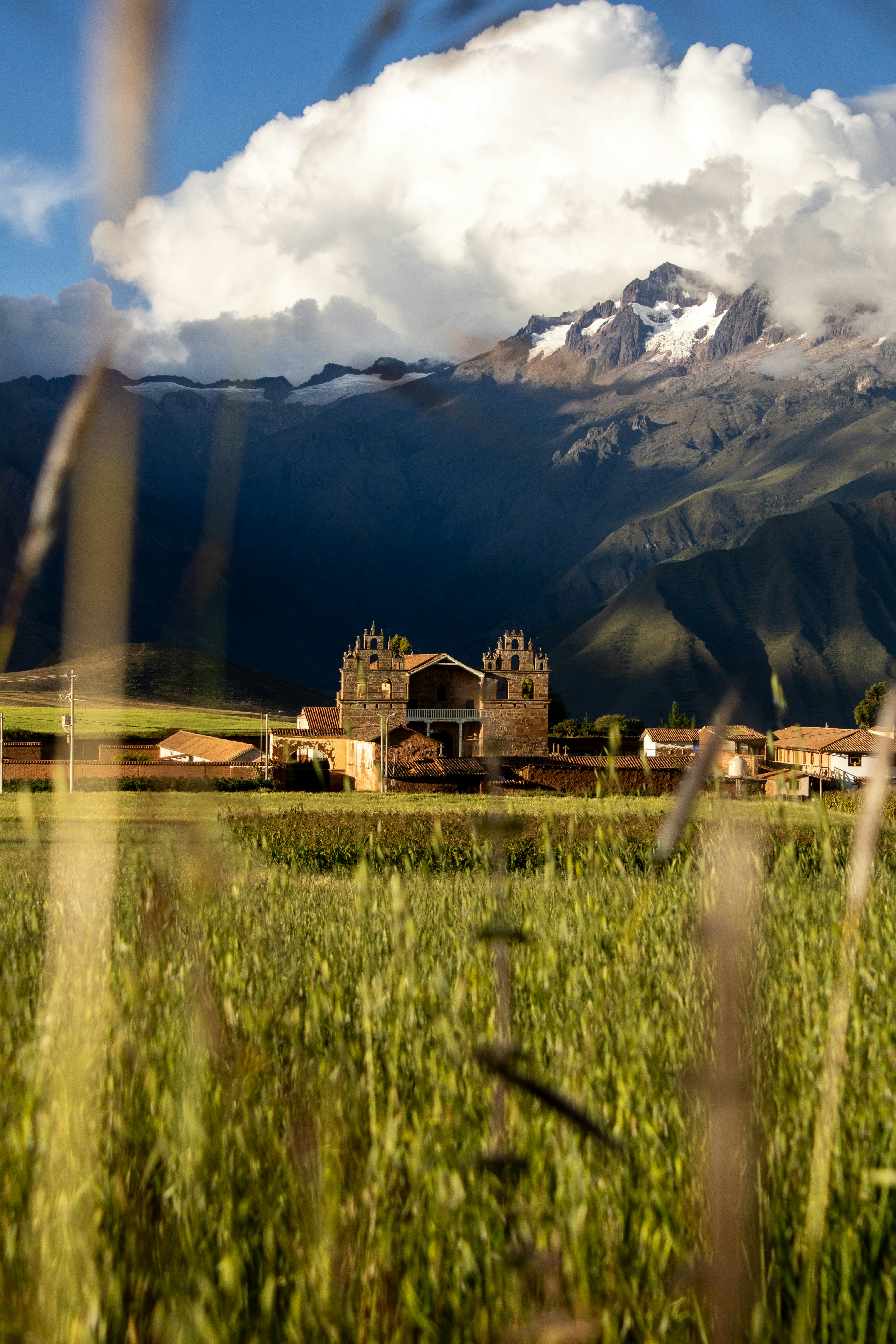 Historic church framed by tall grasses with a backdrop of snow-capped mountains and dramatic clouds.