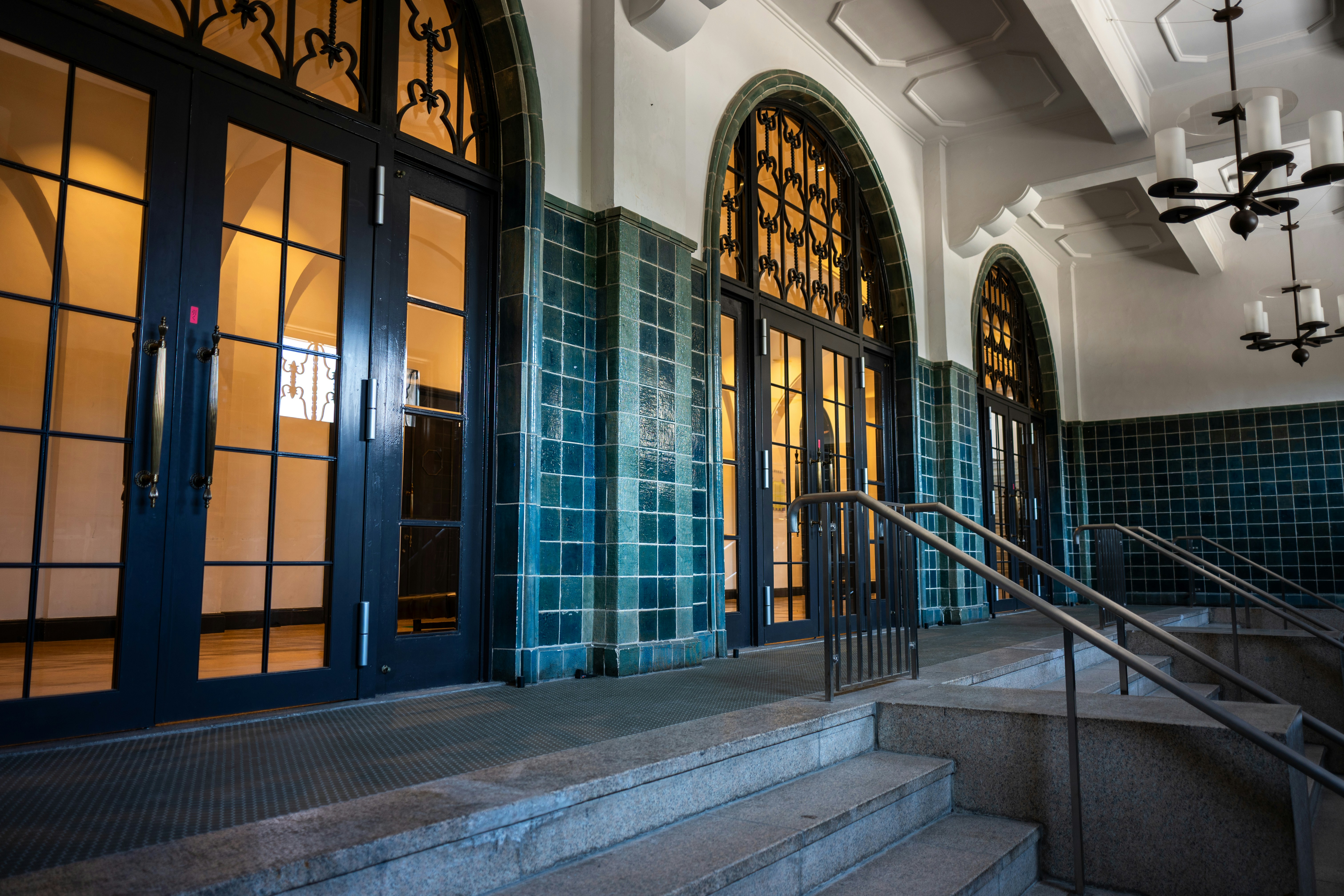 Interior hallway with arched windows, blue-green tiled walls, and geometric light fixtures.