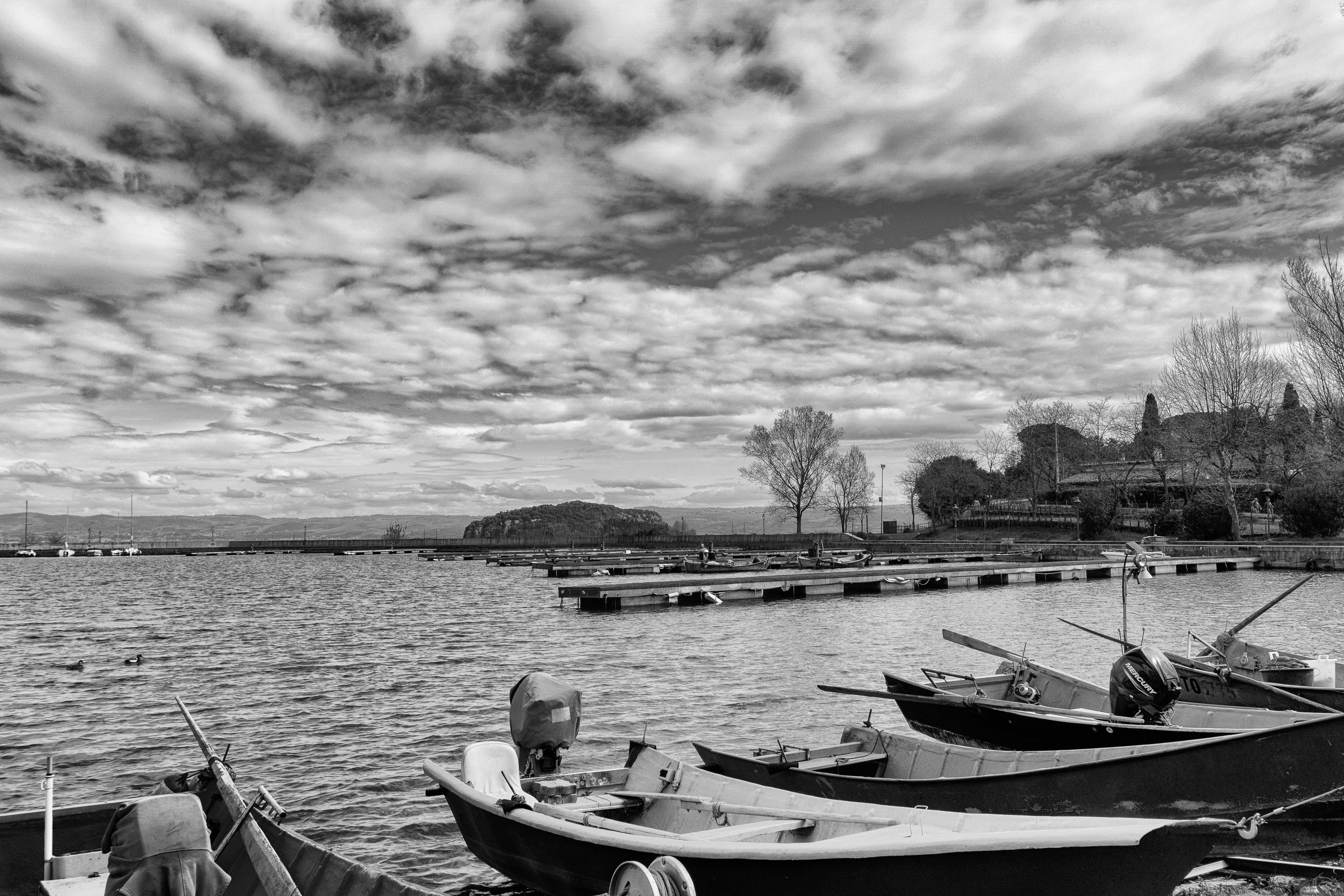 Black and white photograph of boats moored at a dock under a dramatic cloud-filled sky.