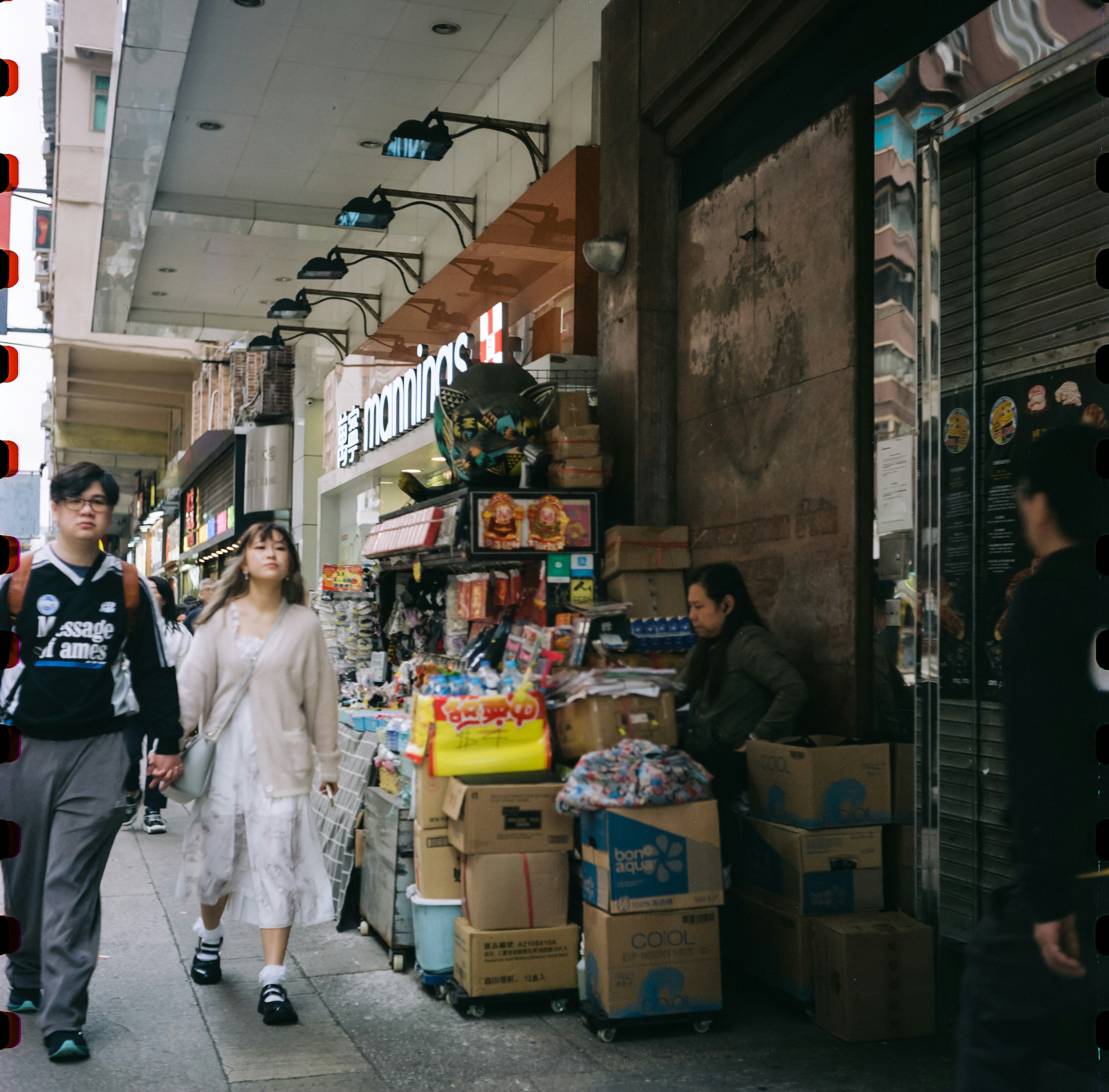 Street vendor sits by a roadside stall with passersby walking past on a busy sidewalk.