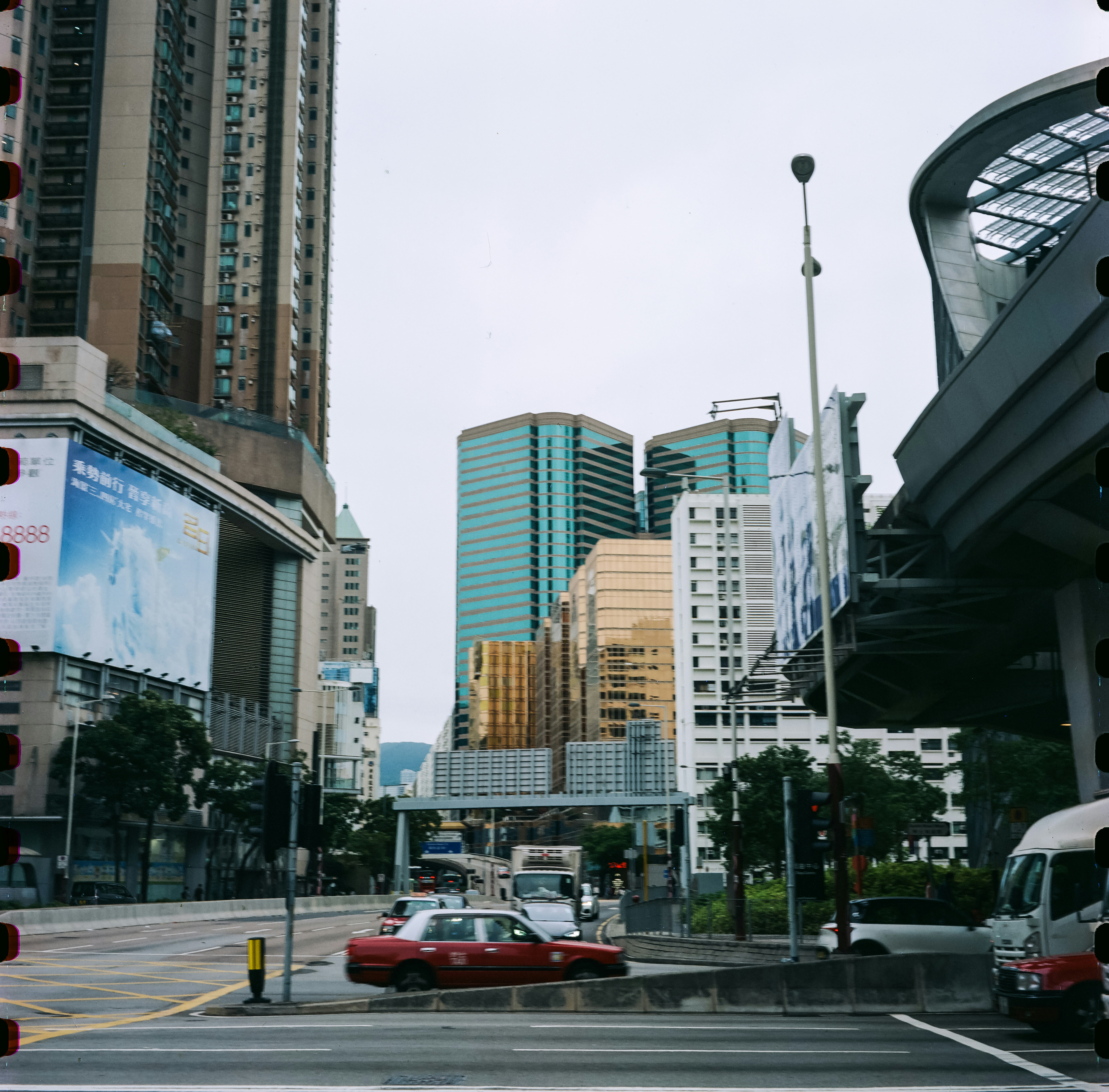 Busy Hong Kong street with skyscrapers, red taxis, and elevated walkways under a cloudy sky.