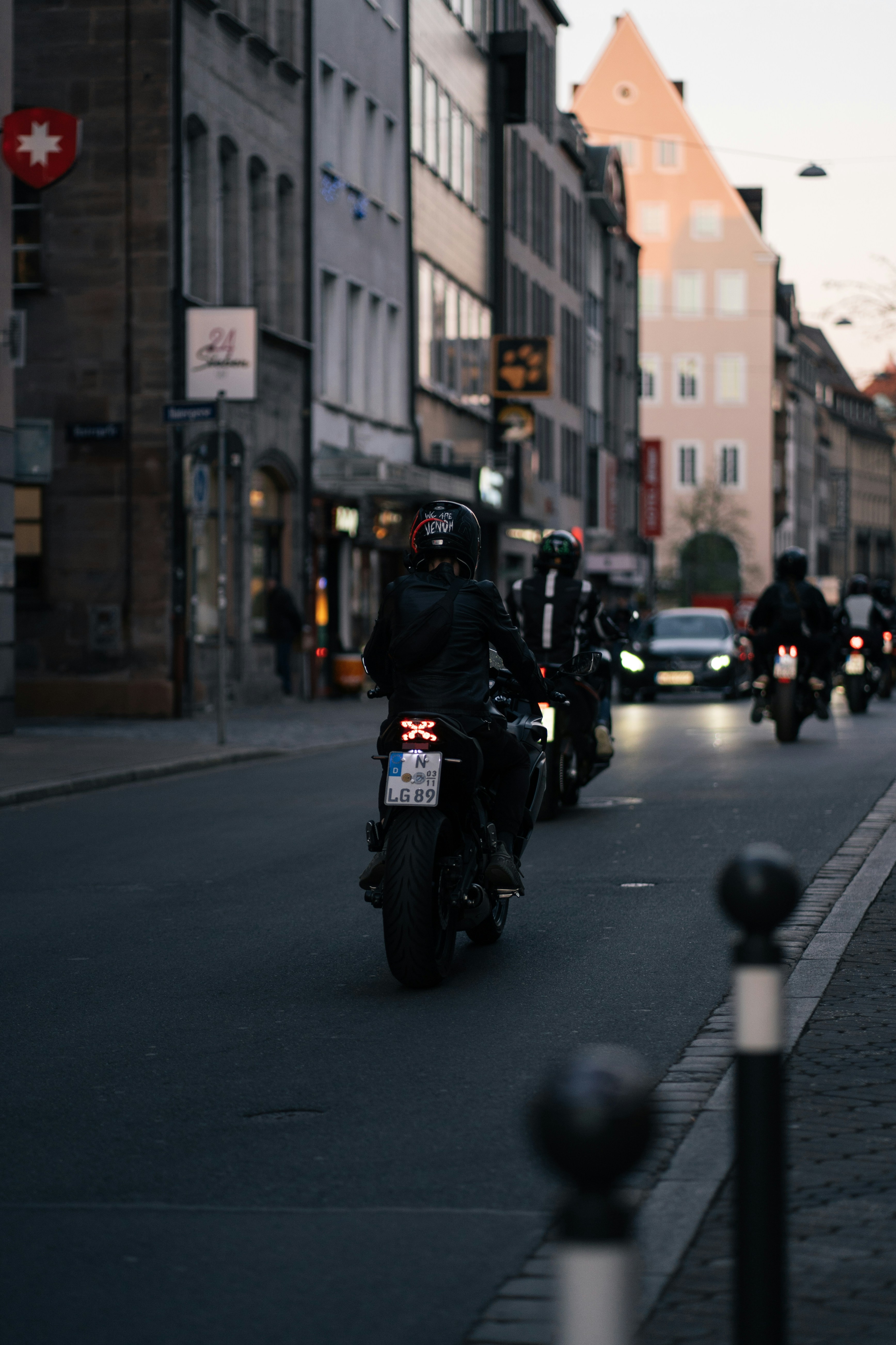 Motorcyclists travel down a dimly lit urban street lined with historic buildings at dusk.