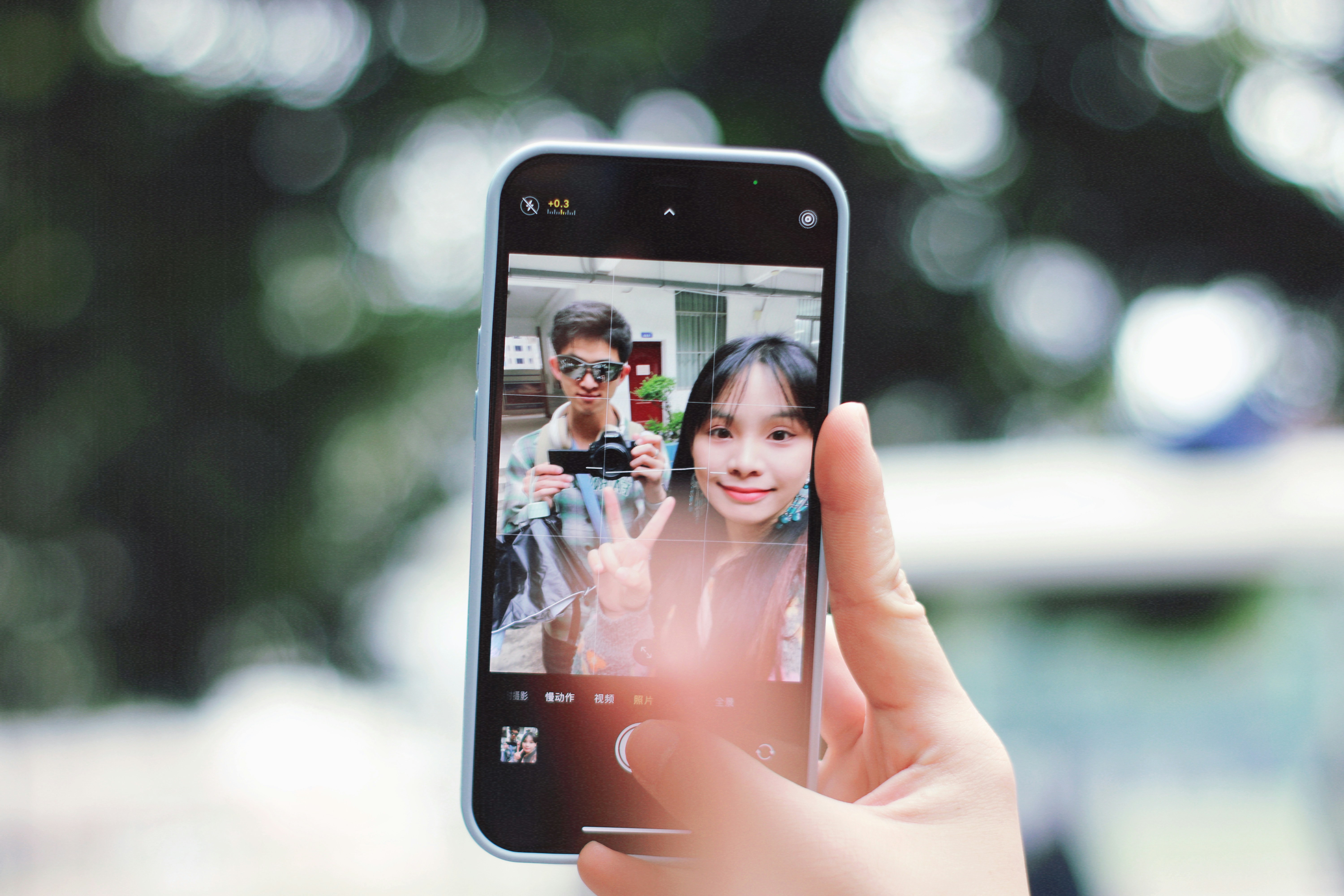 Two people taking a selfie with a smartphone against a blurred natural backdrop.
