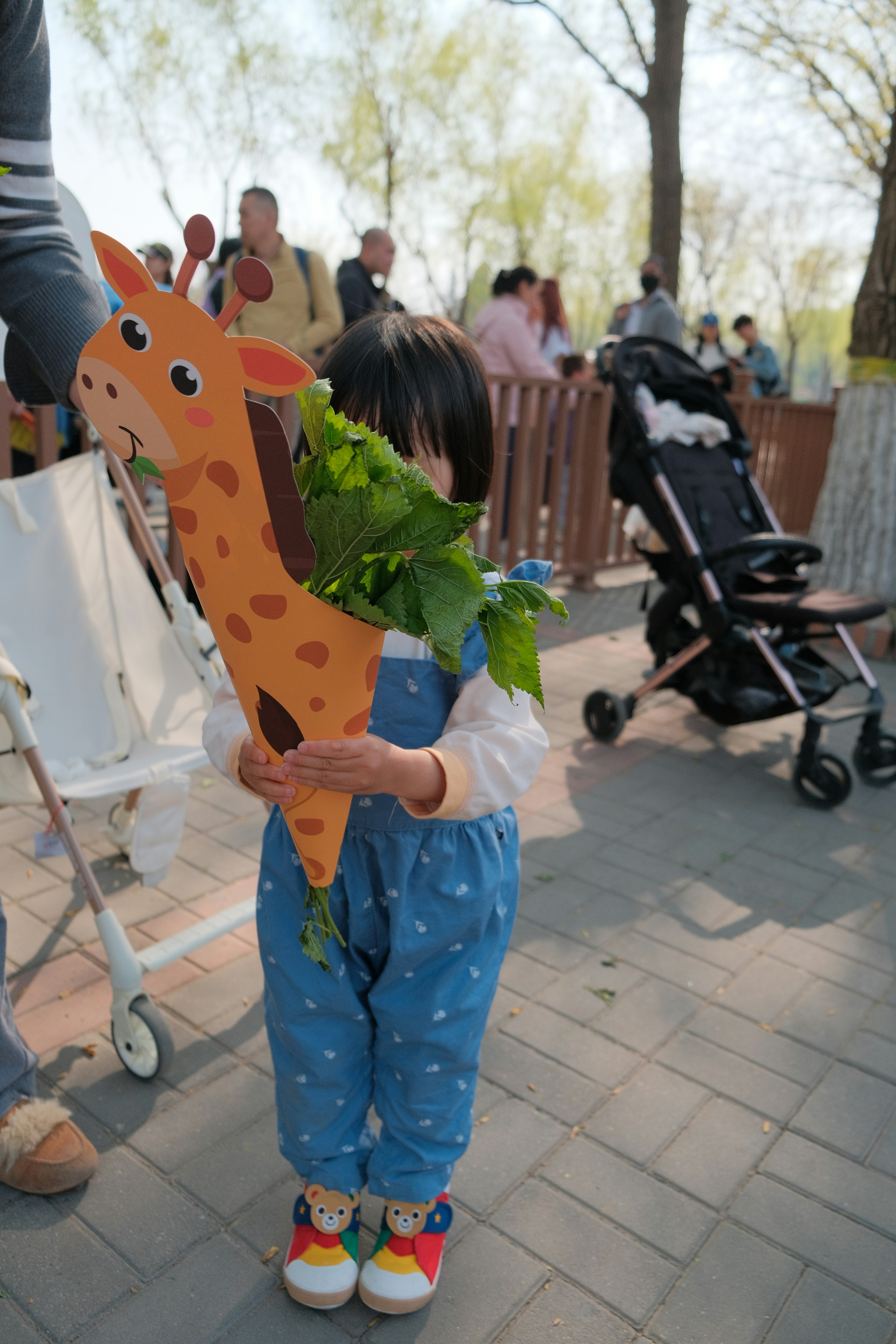 Child holding a giraffe-shaped bouquet against a backdrop of people and strollers in a park.