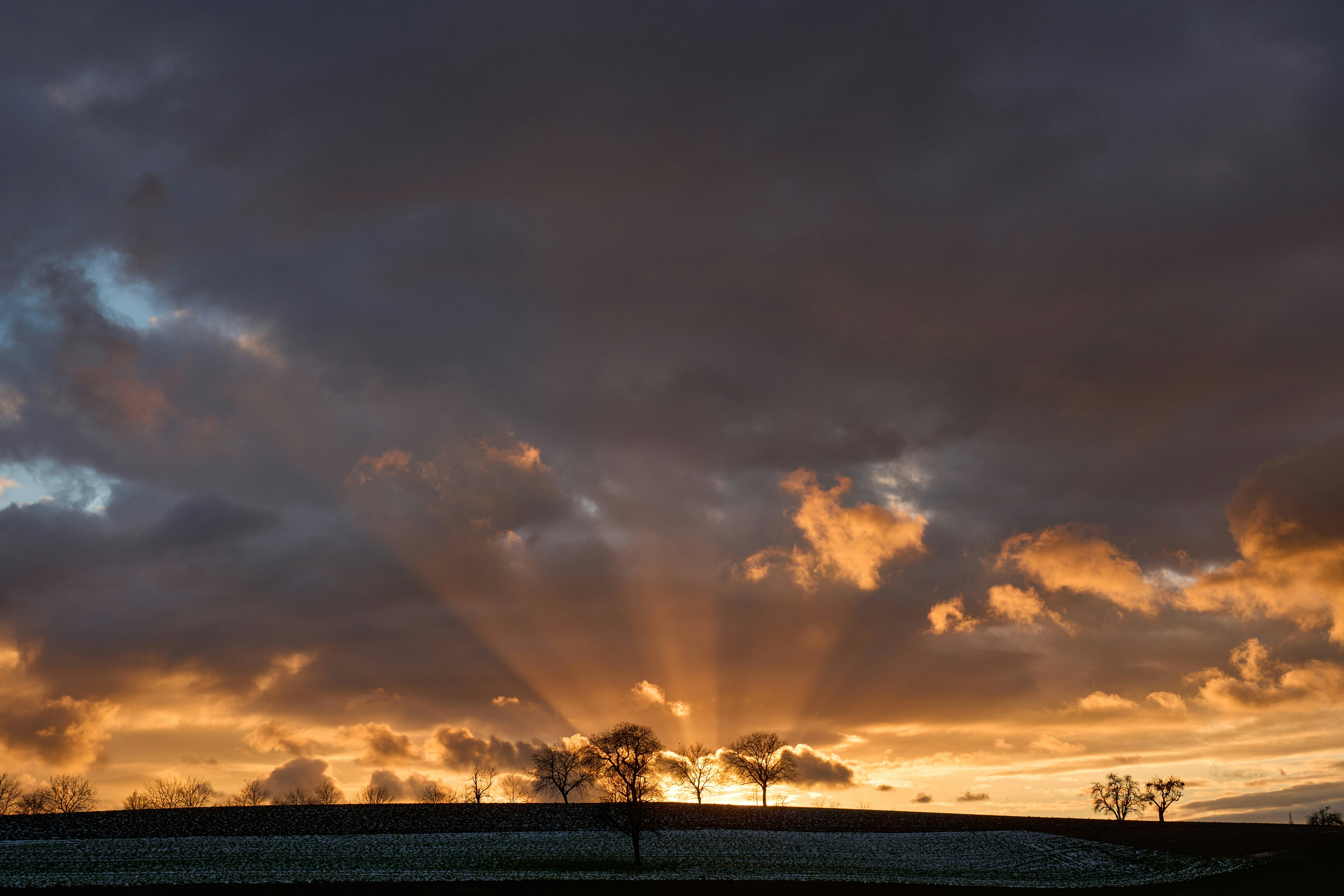 Golden sun rays pierce through dramatic dark clouds at sunset over a silhouetted landscape.